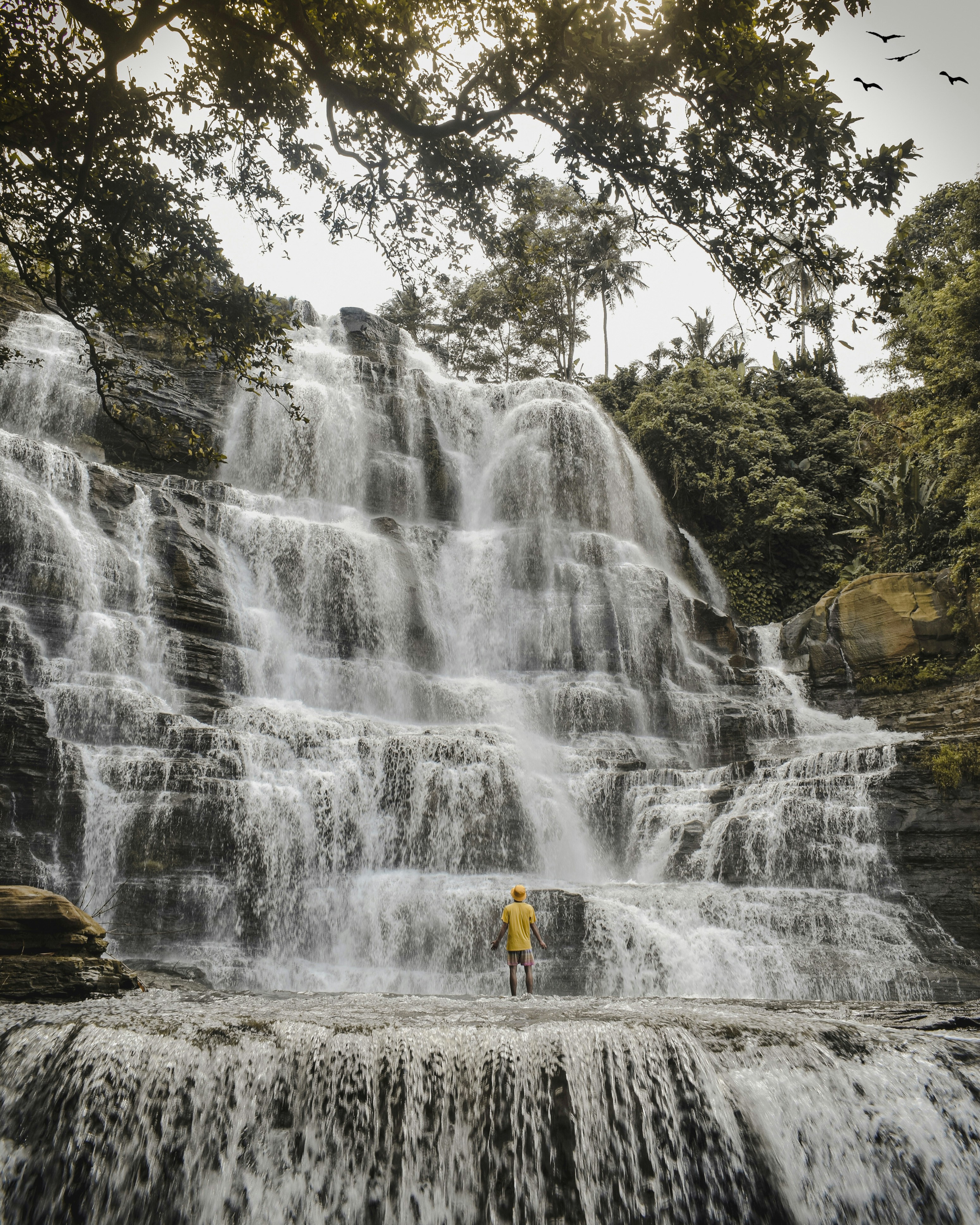 Person in yellow stands at the base of a multi-tiered waterfall surrounded by lush greenery.