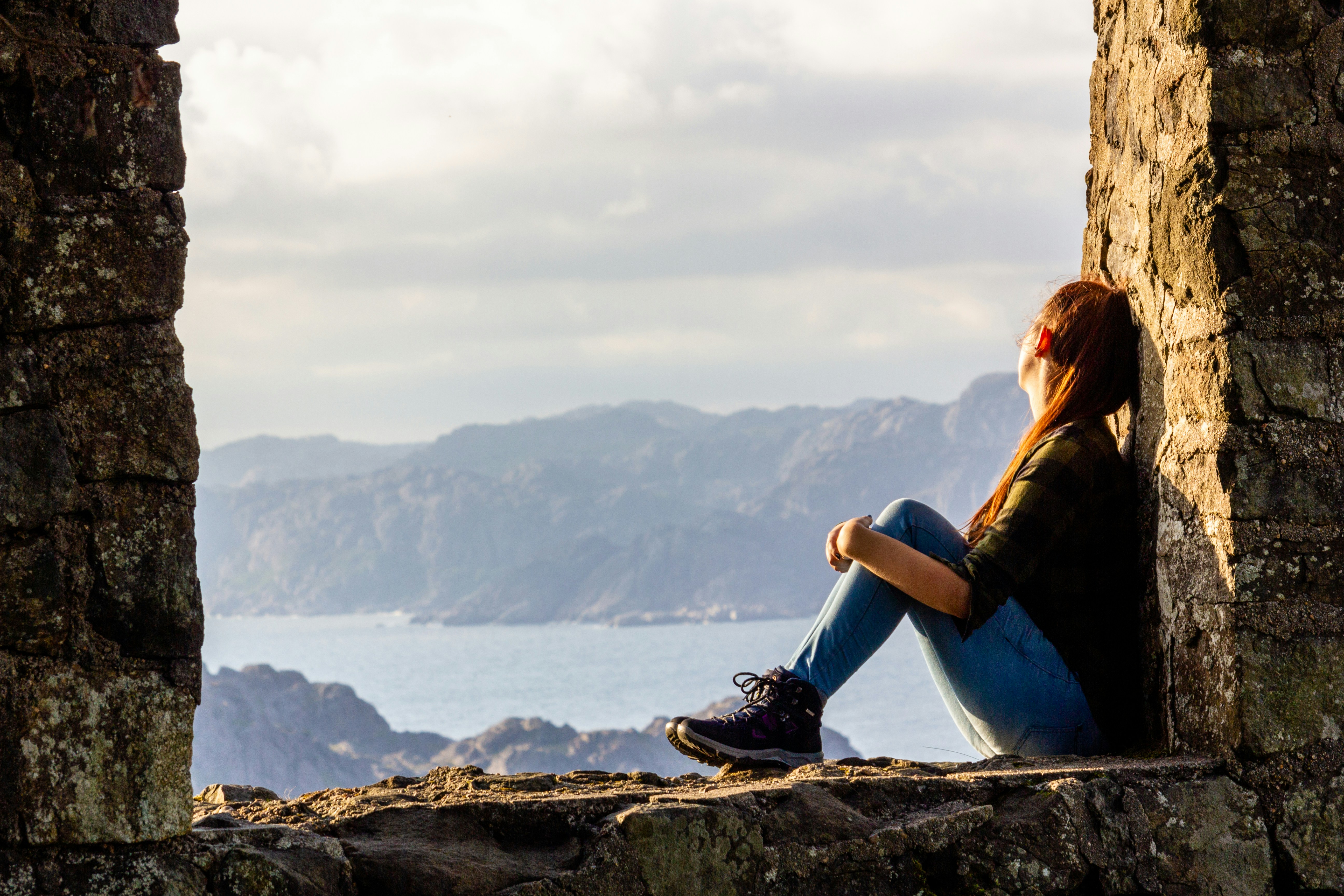 A person sits thoughtfully in a stone window frame, gazing out at a serene seascape with distant mountains. The scene captures a blend of nature and solitude.