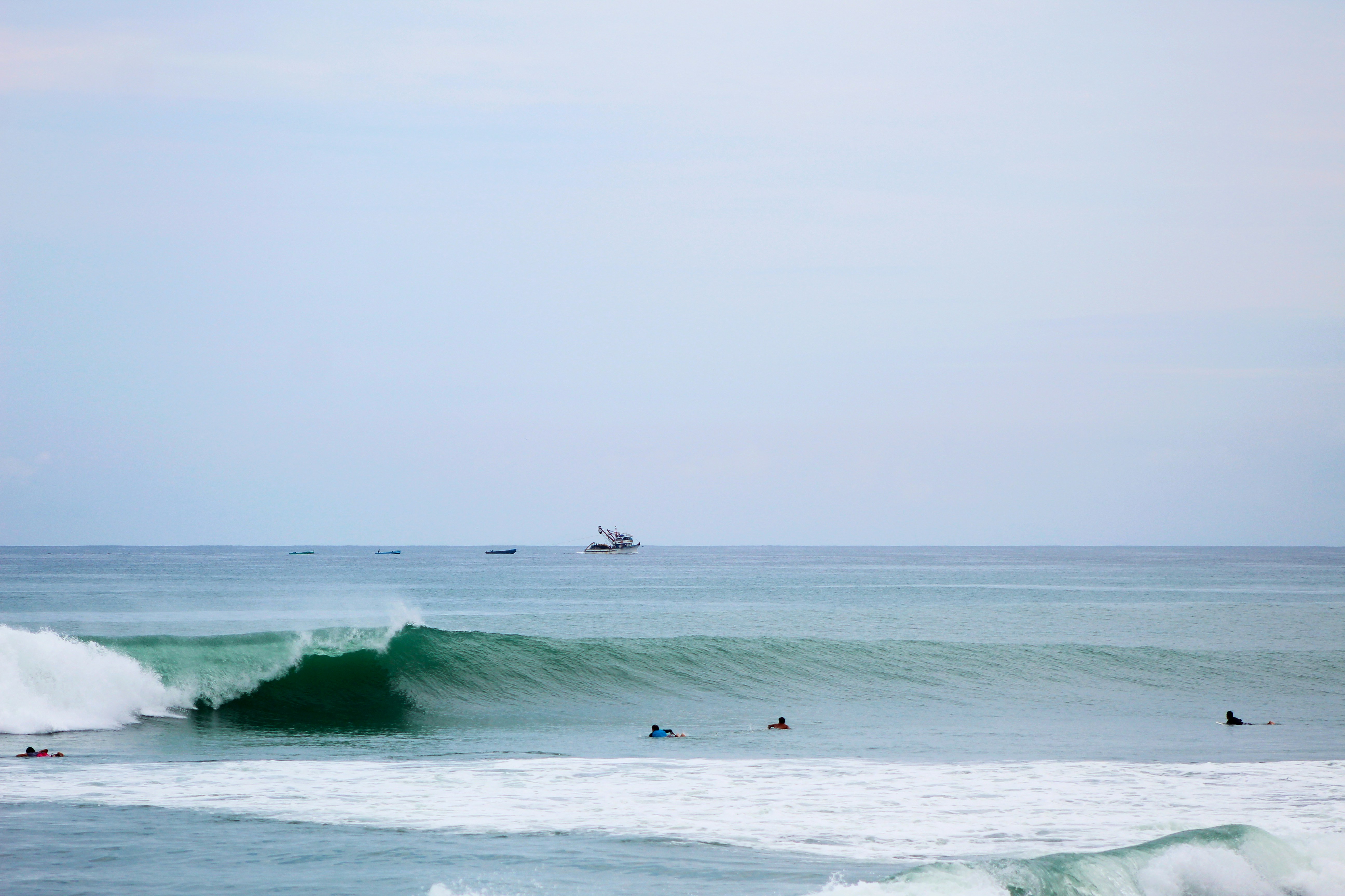 Surfers navigating rolling waves under a cloudy sky with a distant boat on the horizon.