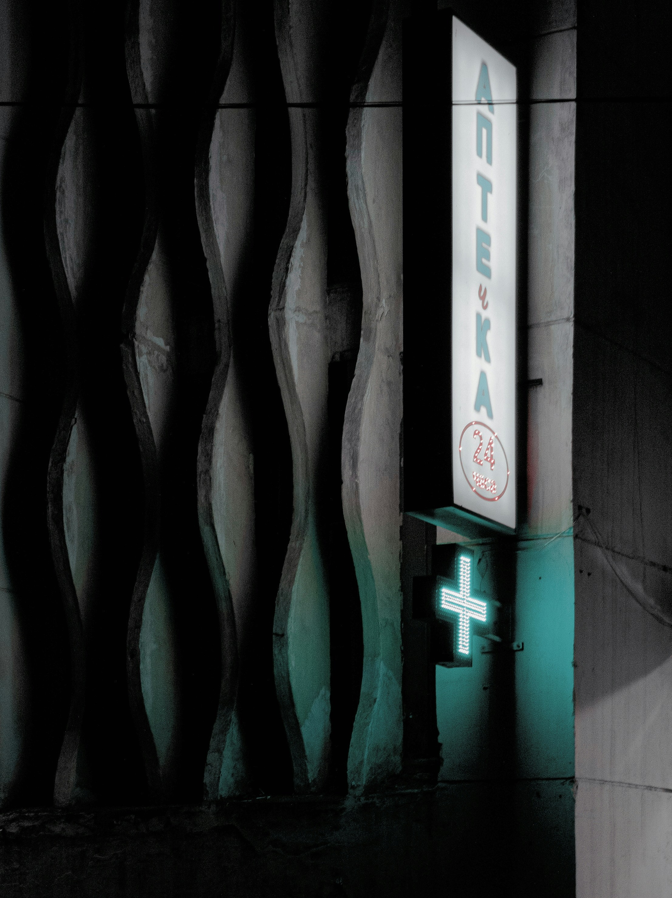 Night photograph of a building facade featuring a vertically illuminated sign and a teal neon cross.