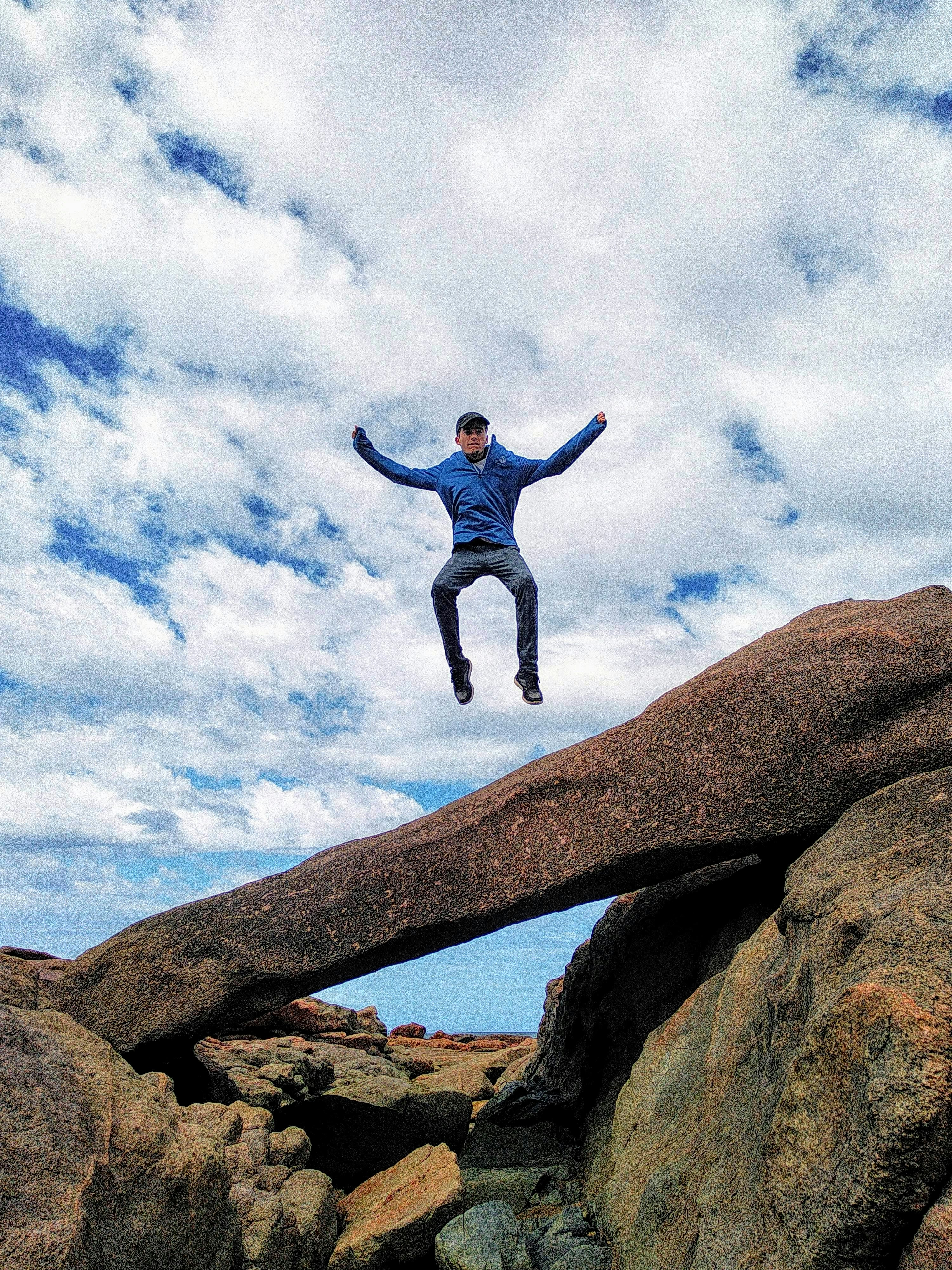 Man leaps mid-air across a rugged granite arch beneath a blue, cloud-filled sky in a landscape photograph.