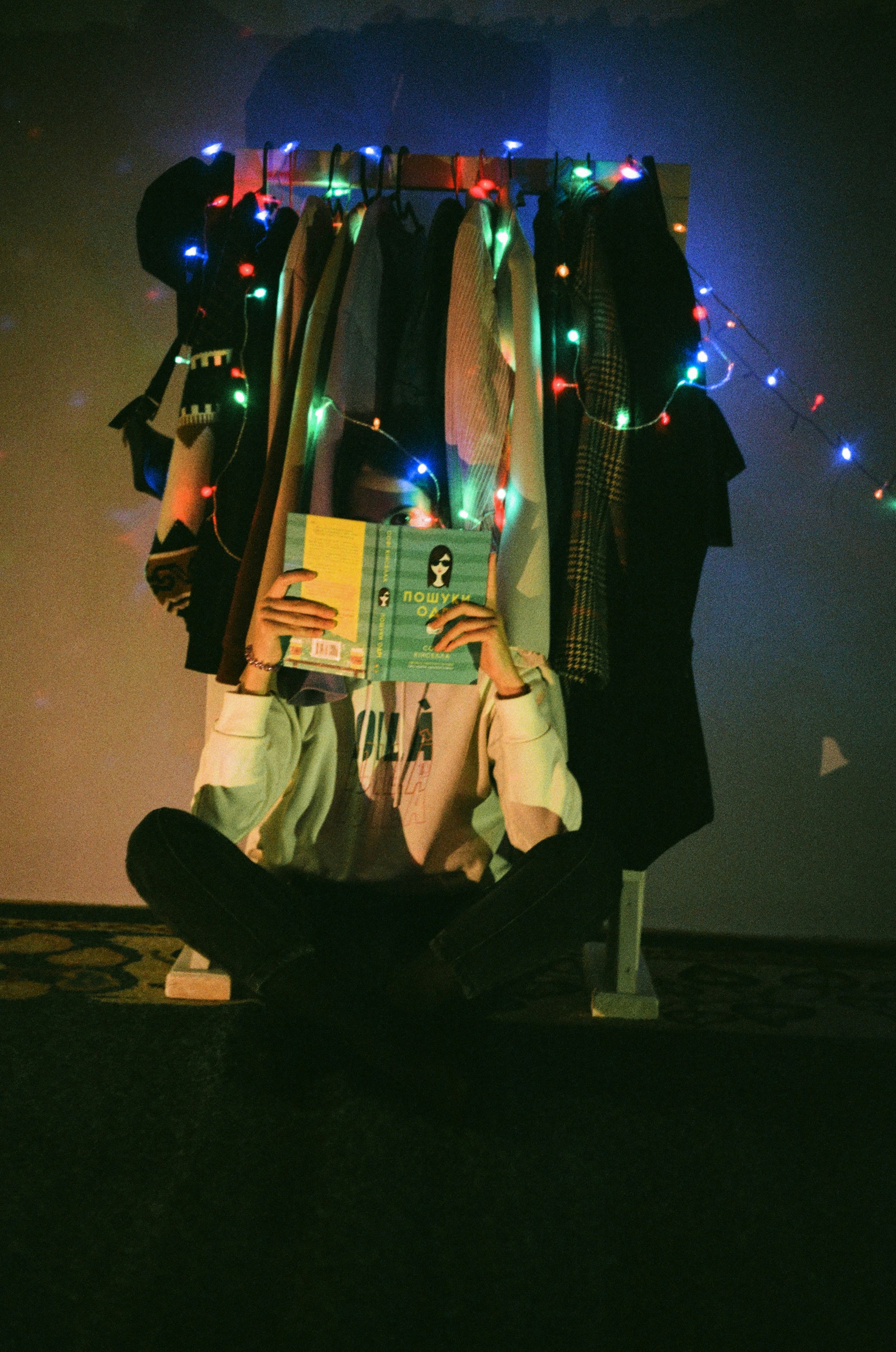 Person seated on the floor, engrossed in a book, surrounded by colorful fairy lights and hanging clothes.
