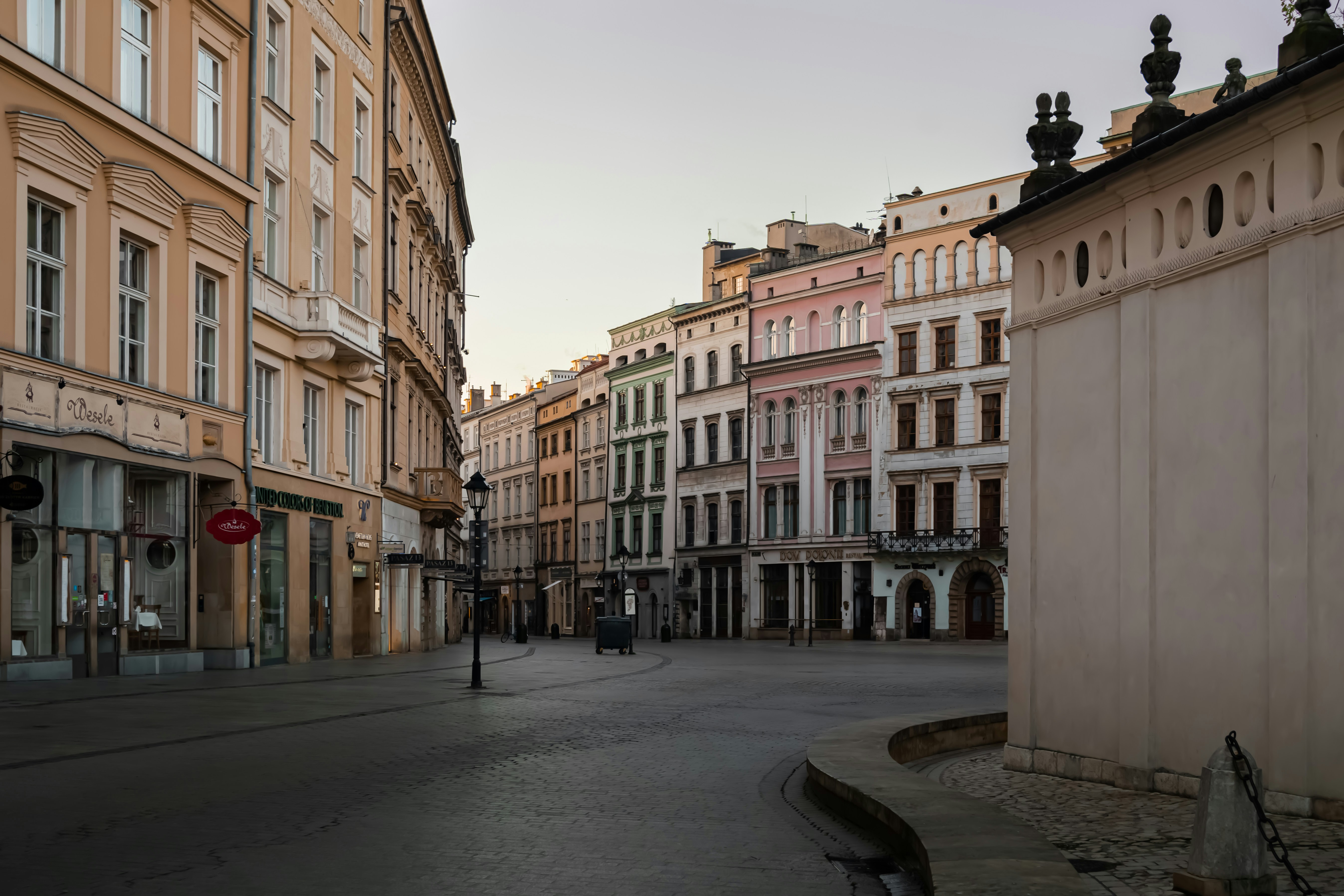 Charming European street lined with pastel-colored buildings, bathed in soft morning light. The scene captures an early hour with an empty cobblestone path.