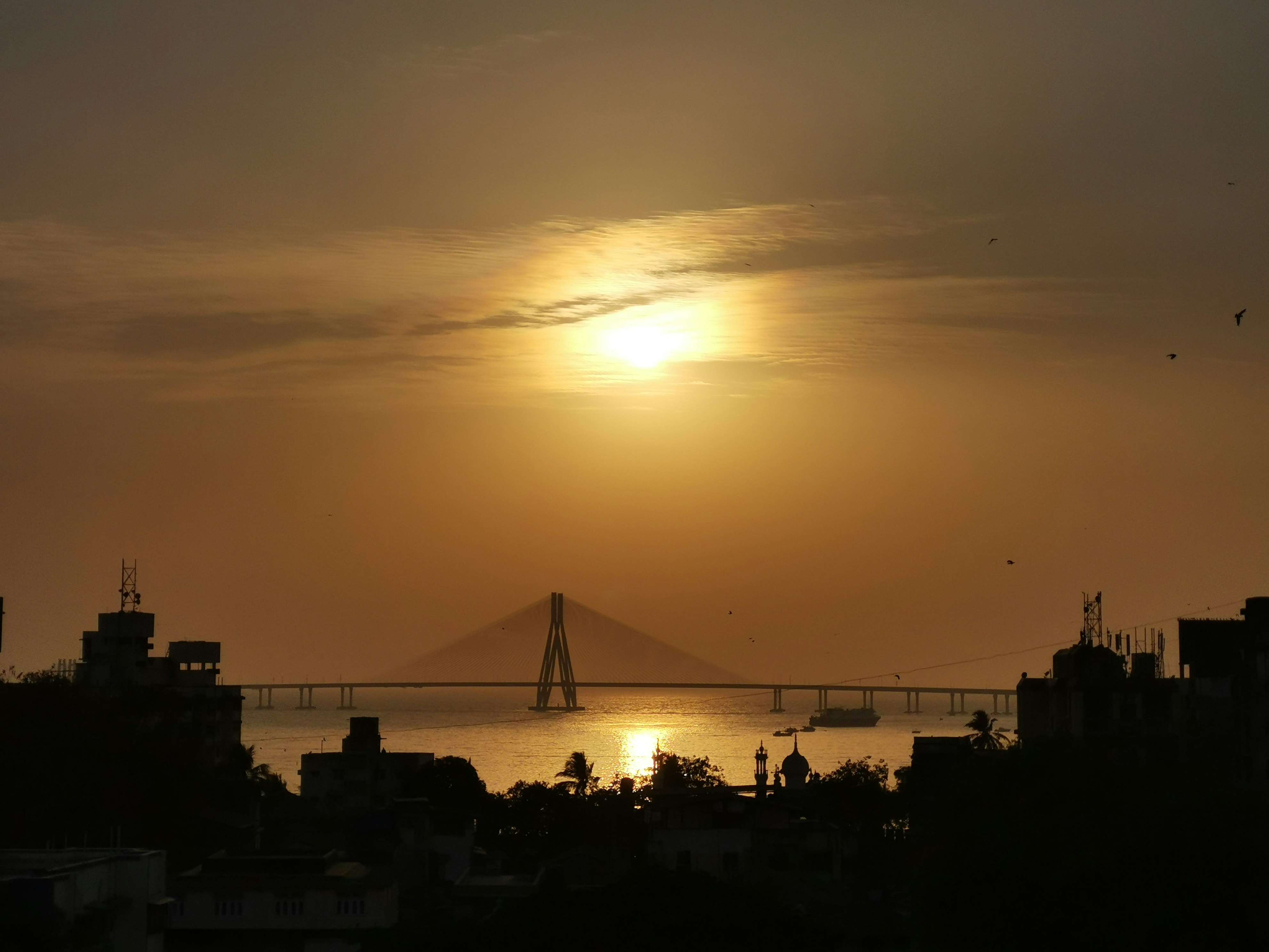 Silhouetted cityscape with a bridge against a warm sunset, casting shadows over the water. The tranquil scene captures the transition from day to night.