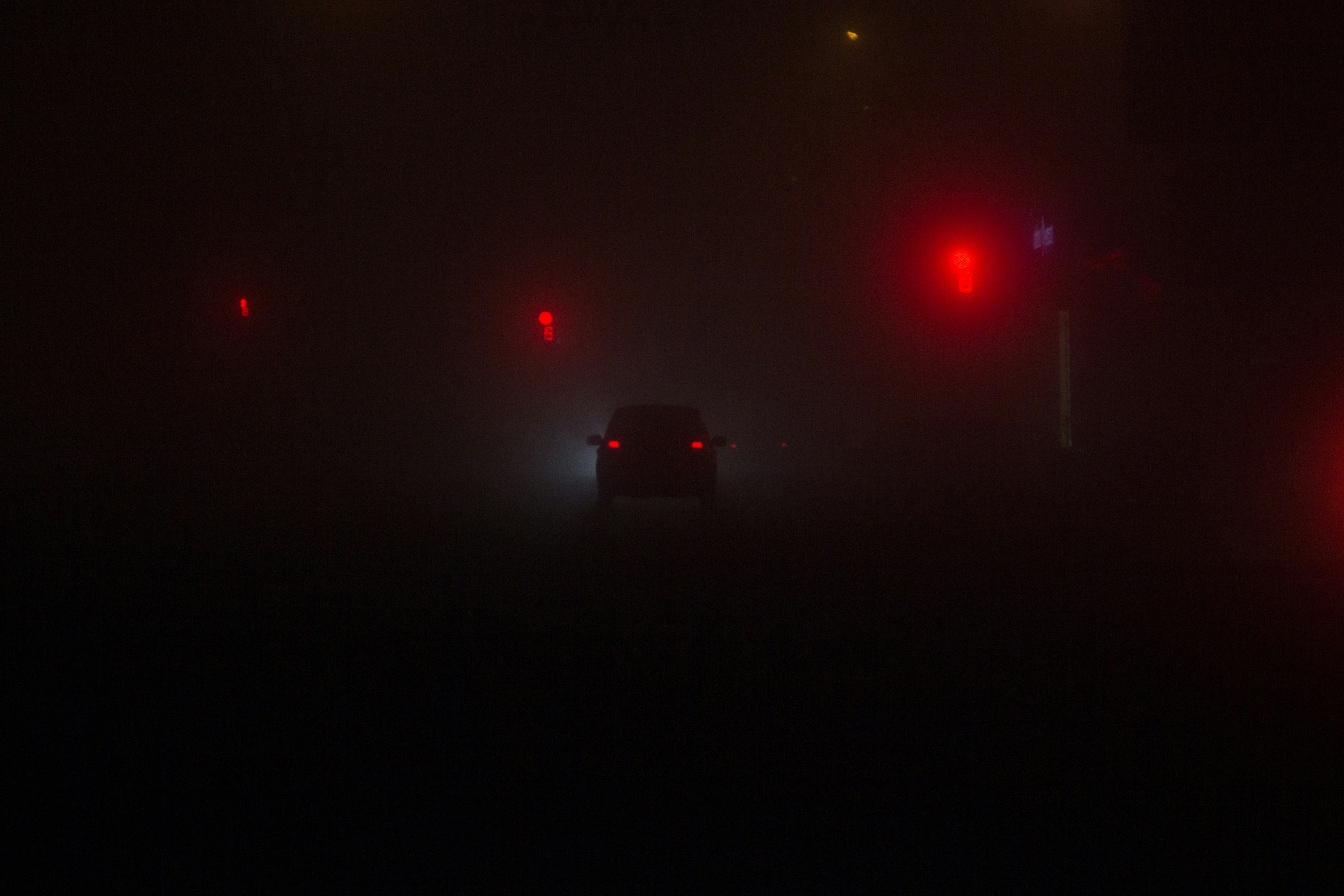 A vehicle navigates through a dense fog, illuminated by distant traffic lights. The scene conveys a sense of mystery and isolation.