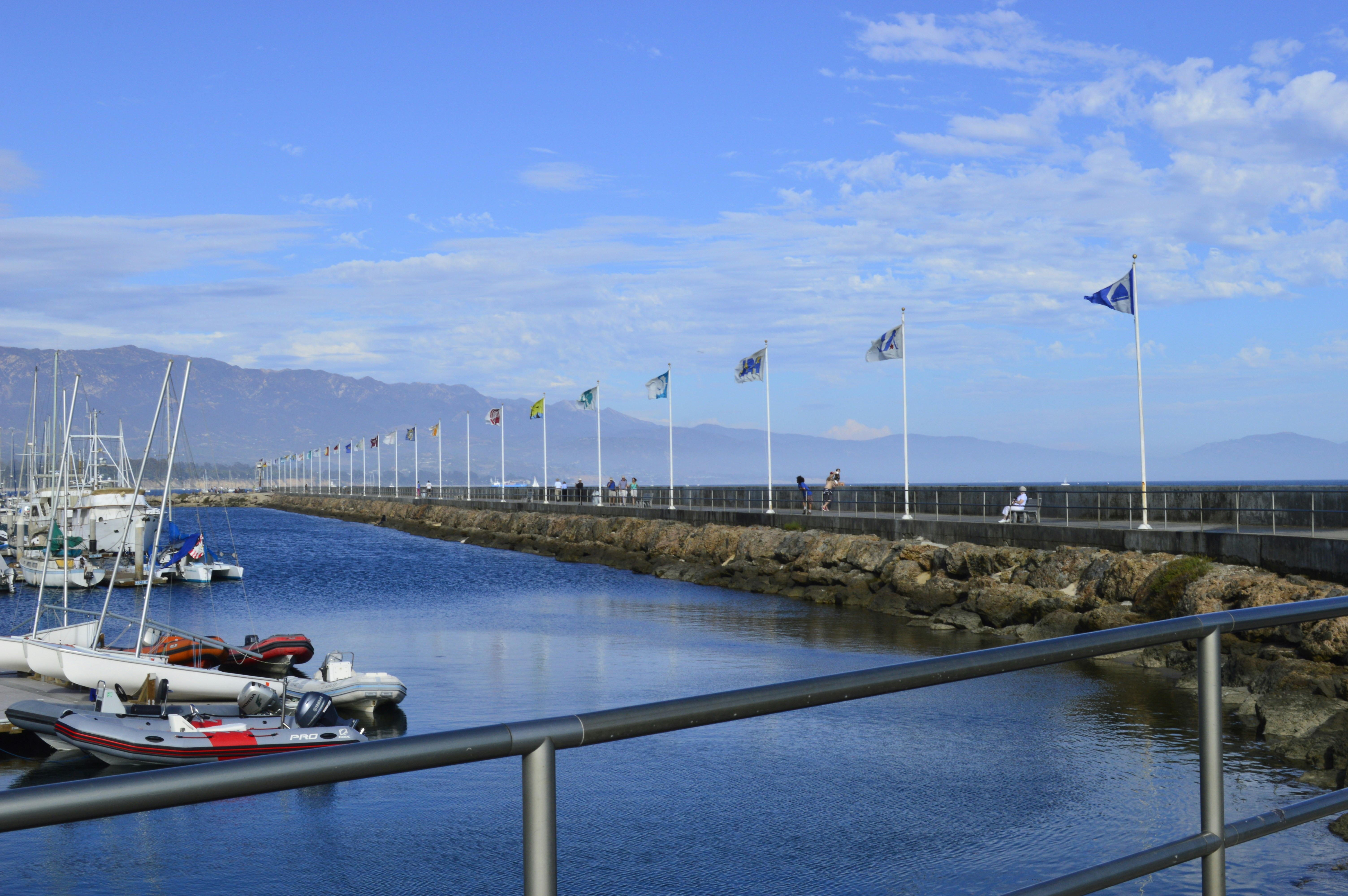 A scenic harbor view featuring moored boats and a walkway lined with flags, set against a backdrop of distant mountains and a clear blue sky.