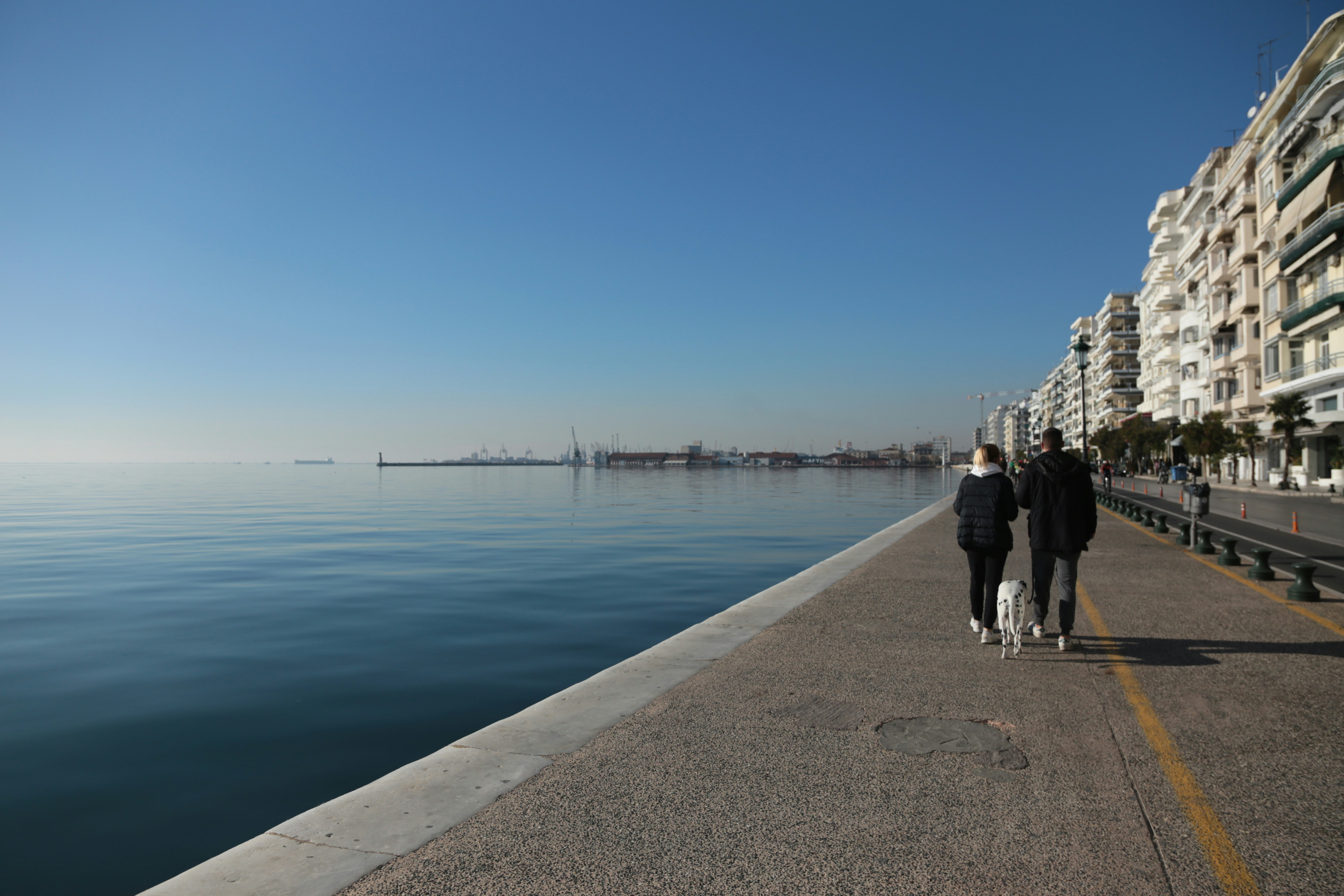 Two figures walking along a smooth waterfront promenade under a clear sky, with calm waters reflecting the horizon. The urban landscape features modern buildings lining the path.