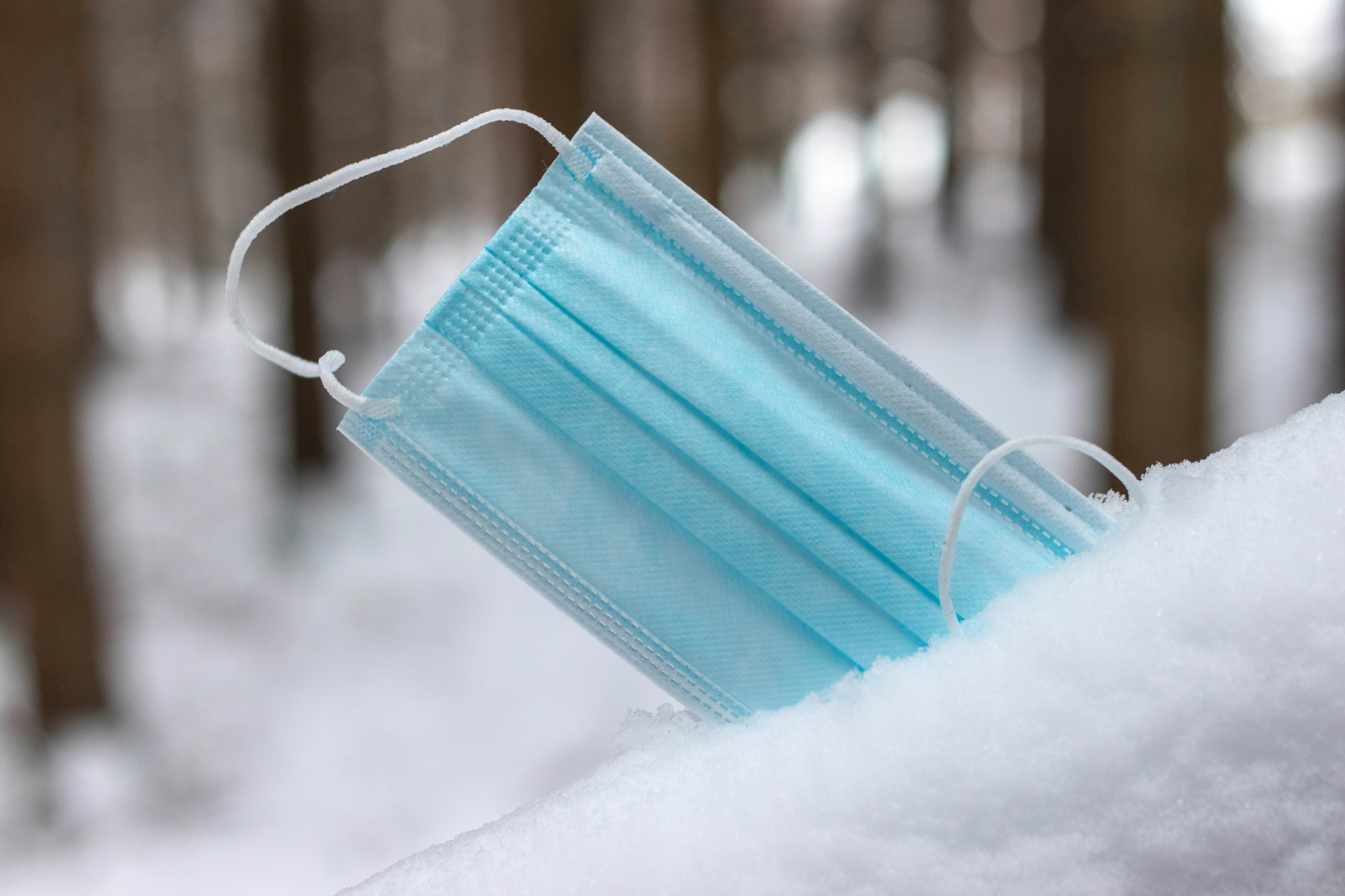 A blue surgical mask partially buried in fresh snow, surrounded by blurred trees in a serene winter setting.