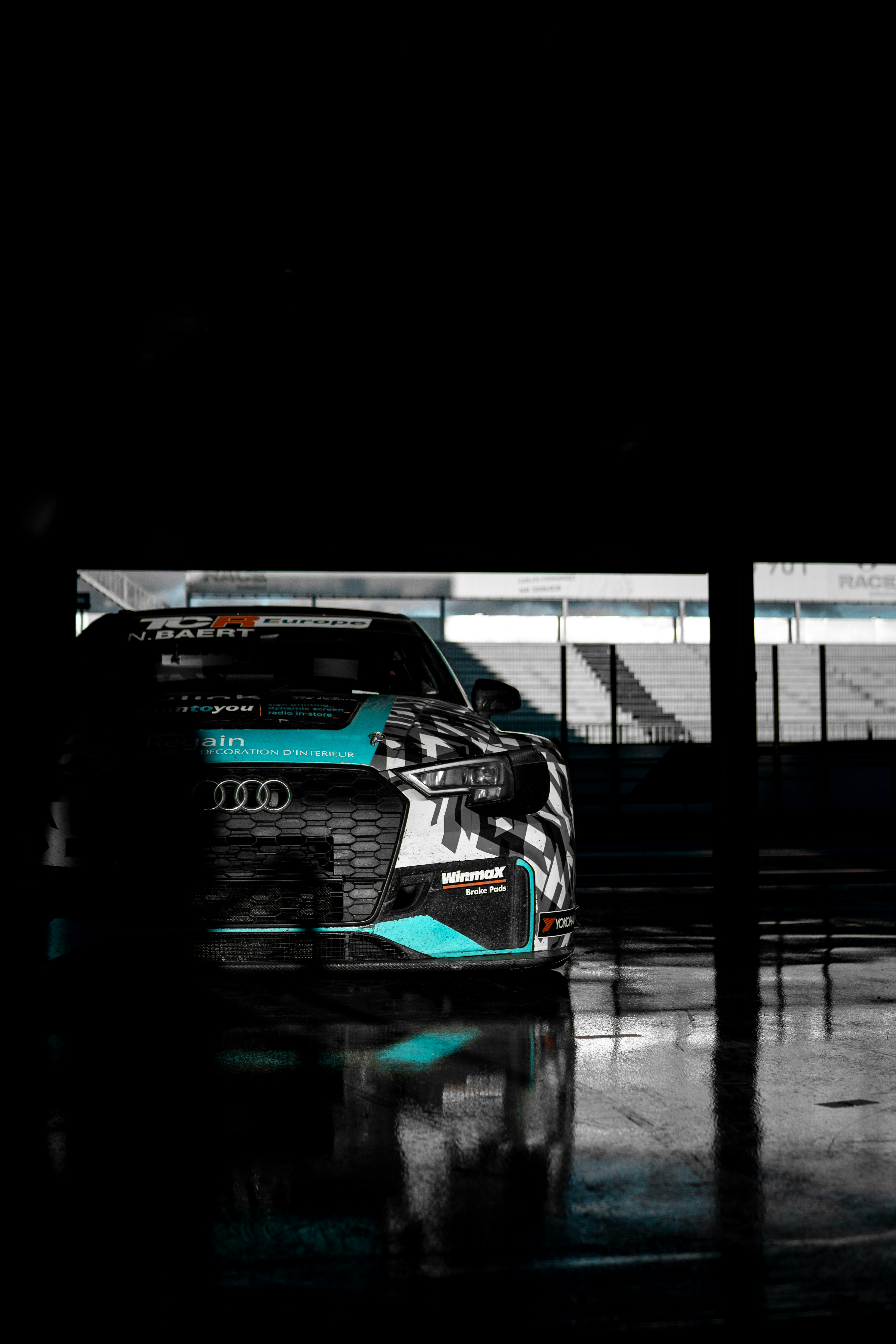 An Audi race car partially illuminated under a structure, showcasing its sleek design and vibrant colors against a dark backdrop. The reflective surface adds depth to the scene.