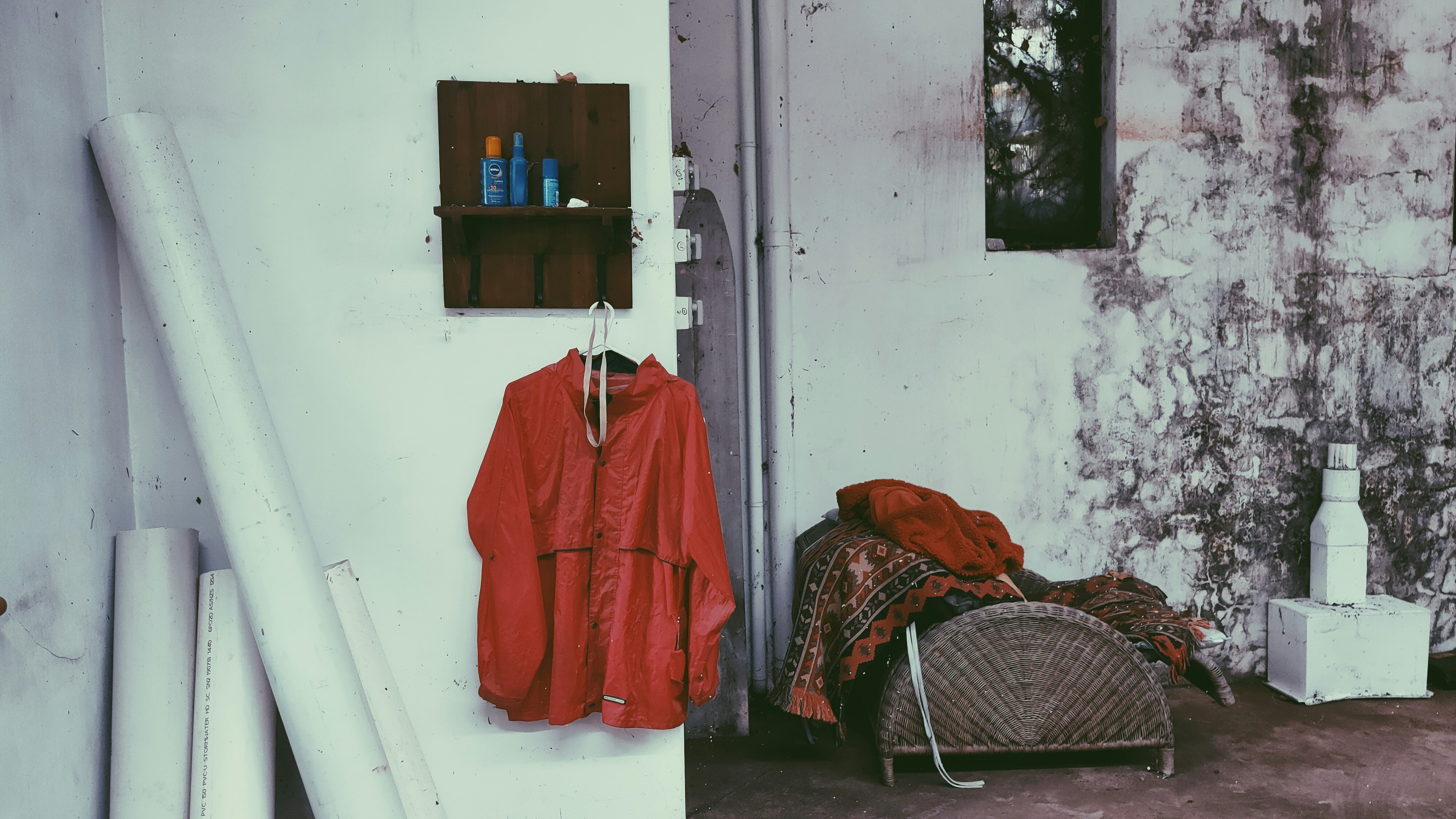 A vibrant red raincoat hangs on a wall beside a wooden shelf holding blue bottles, set against a backdrop of peeling paint and a wicker chair draped with fabric.