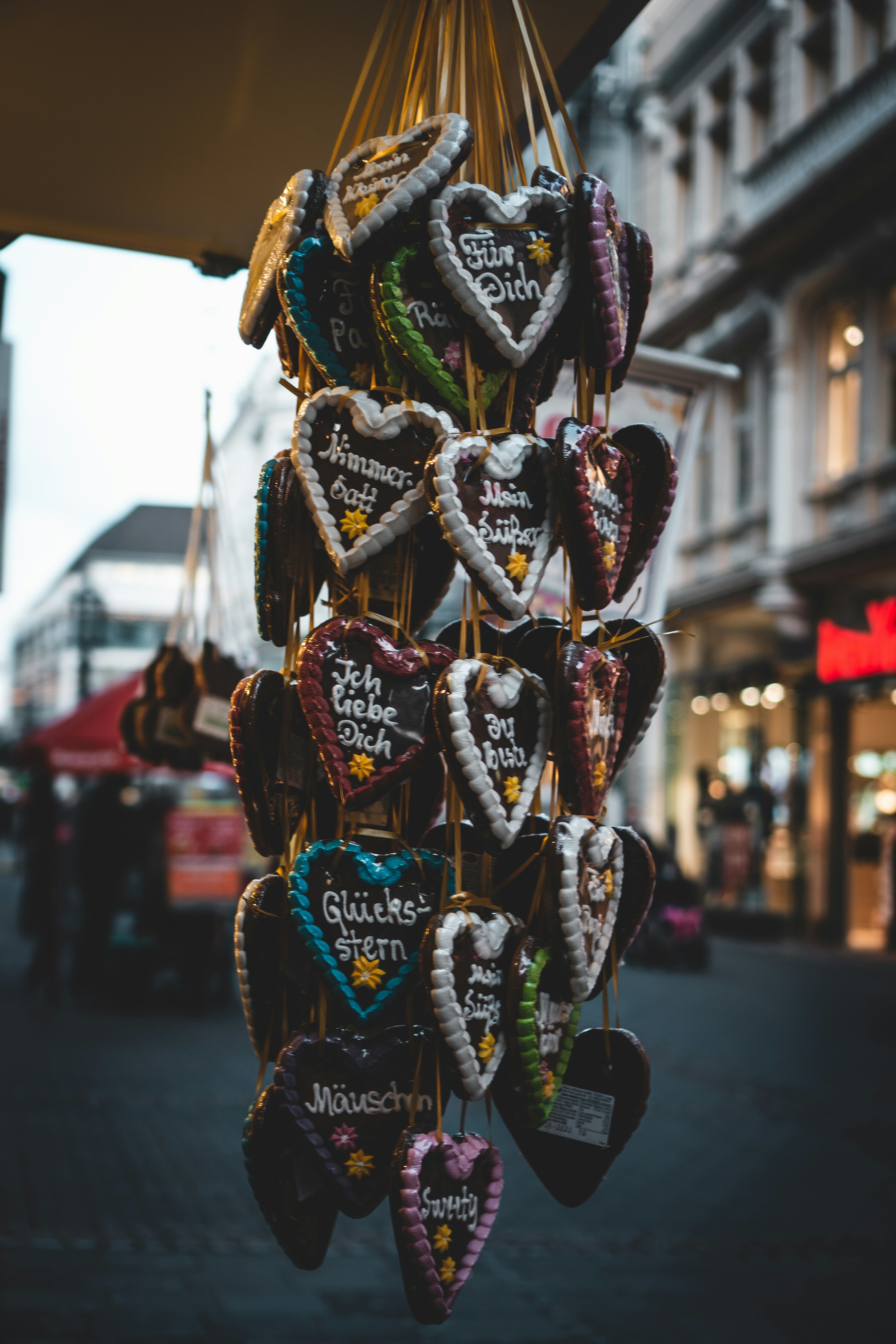 Colorful gingerbread hearts hanging in a bustling market, adorned with playful messages. The scene captures a festive atmosphere filled with charm.