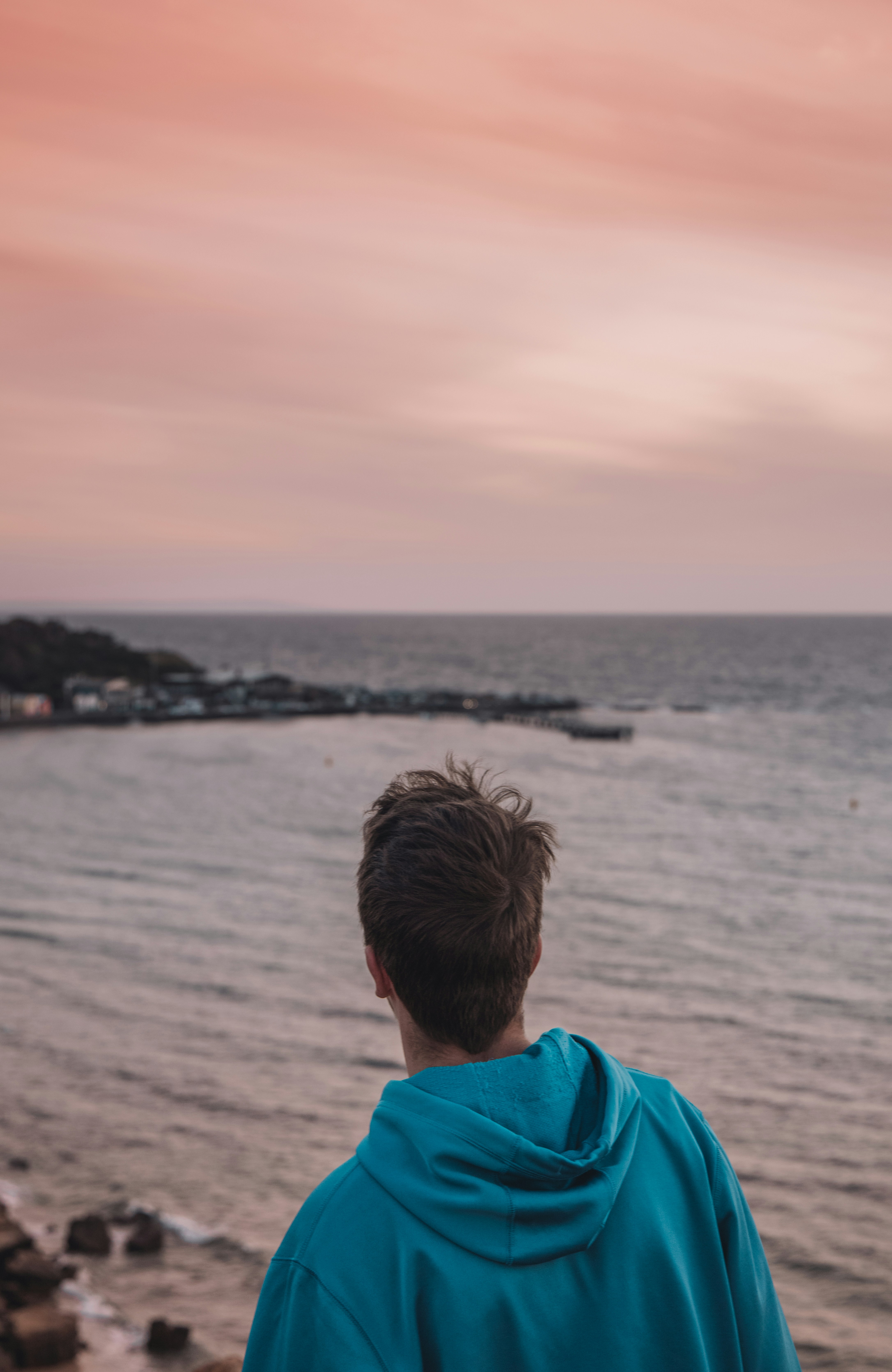 A person in a blue hoodie gazes towards the ocean, with a colorful sky at dusk. The serene coastline adds to the contemplative atmosphere.