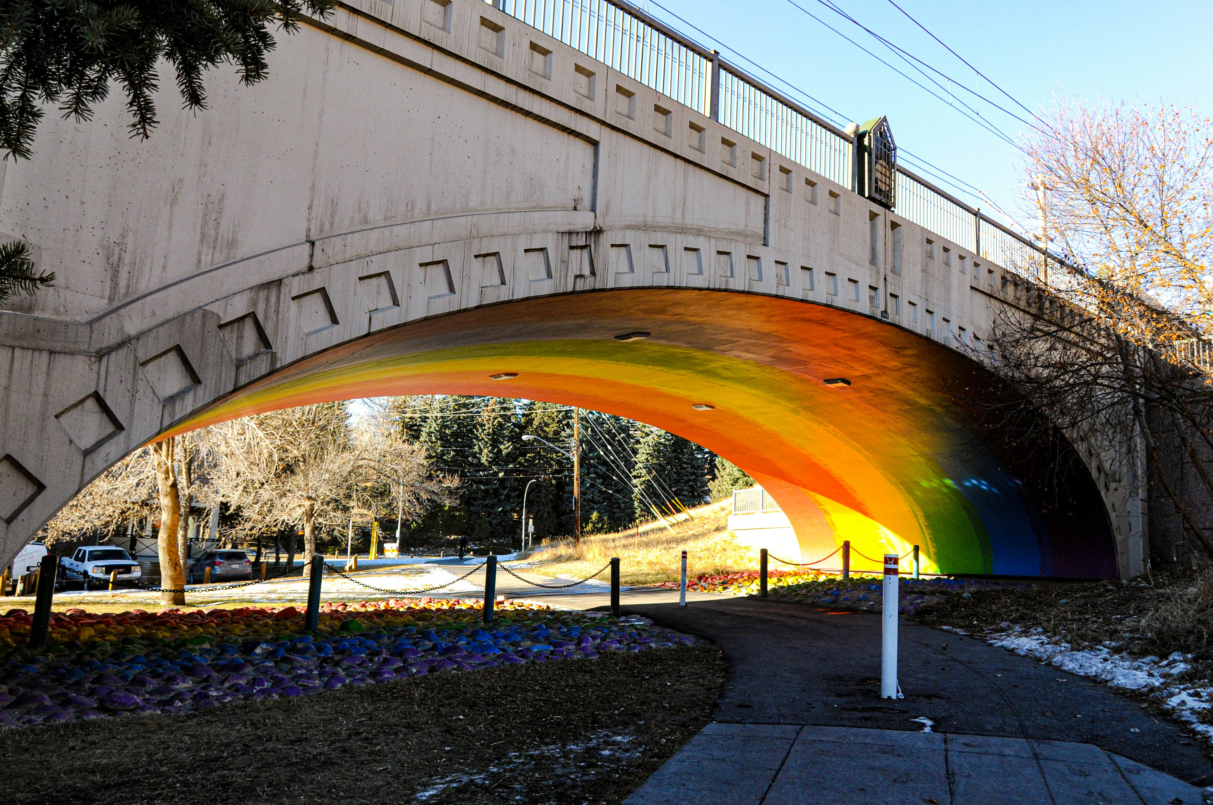 Colorful rainbow mural painted under a bridge, surrounded by trees and a pathway, creating a lively urban atmosphere.