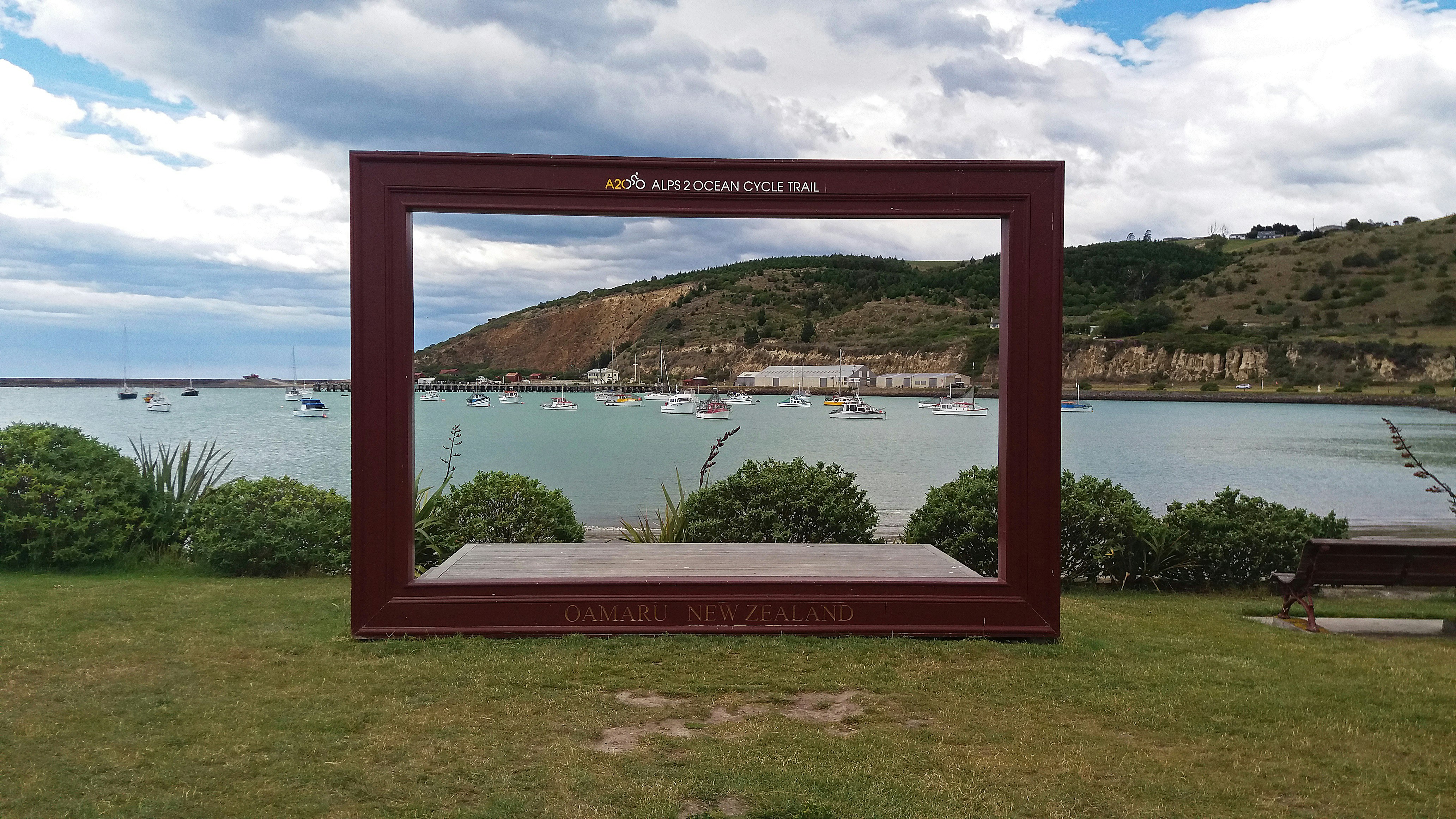 A large, rectangular frame stands in a grassy area, overlooking a calm bay dotted with boats and framed by rolling hills. The scene captures the tranquil essence of Oamaru, New Zealand.