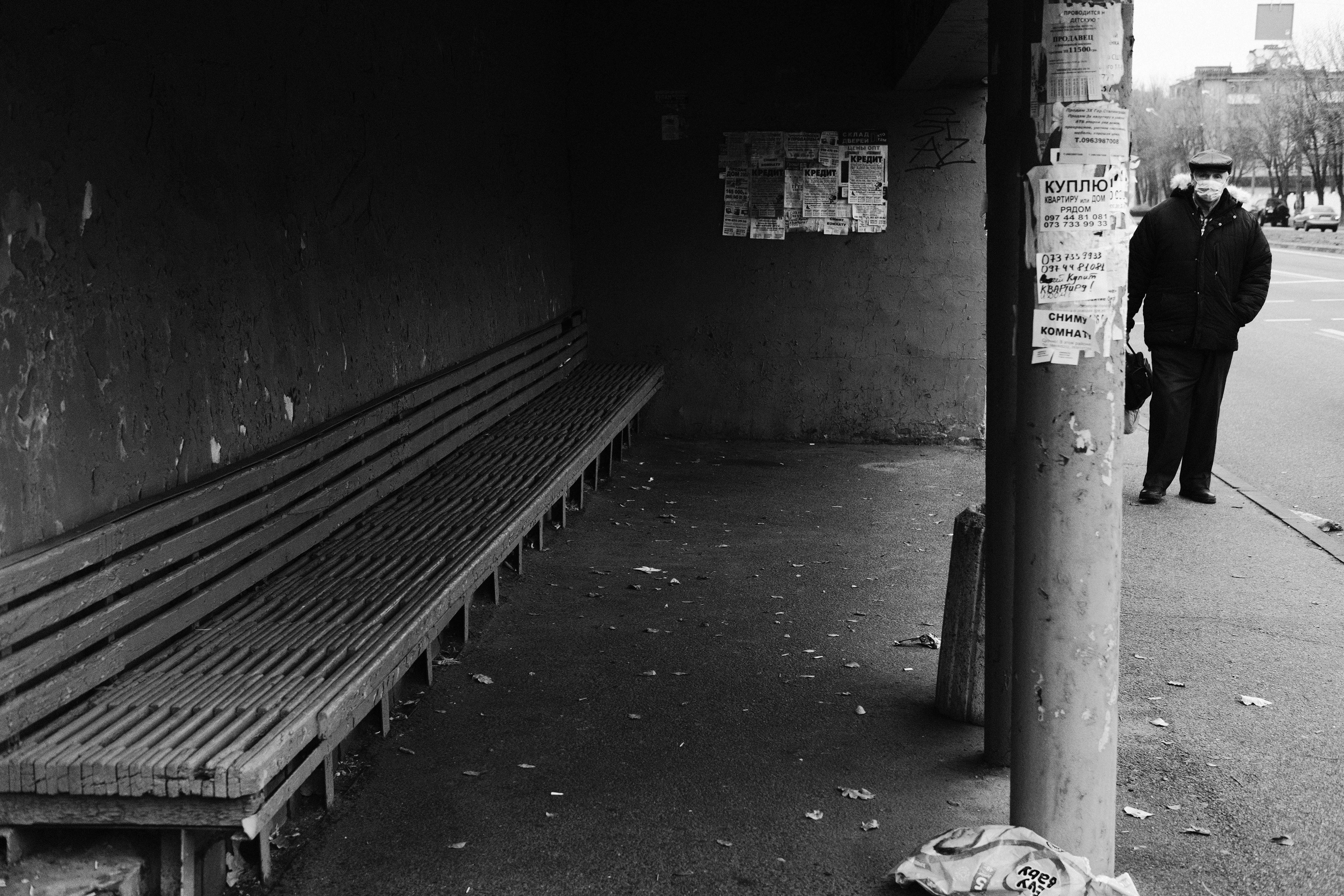 A solitary figure stands near a weathered bus stop, surrounded by posters and an empty bench, evoking a sense of waiting and contemplation.