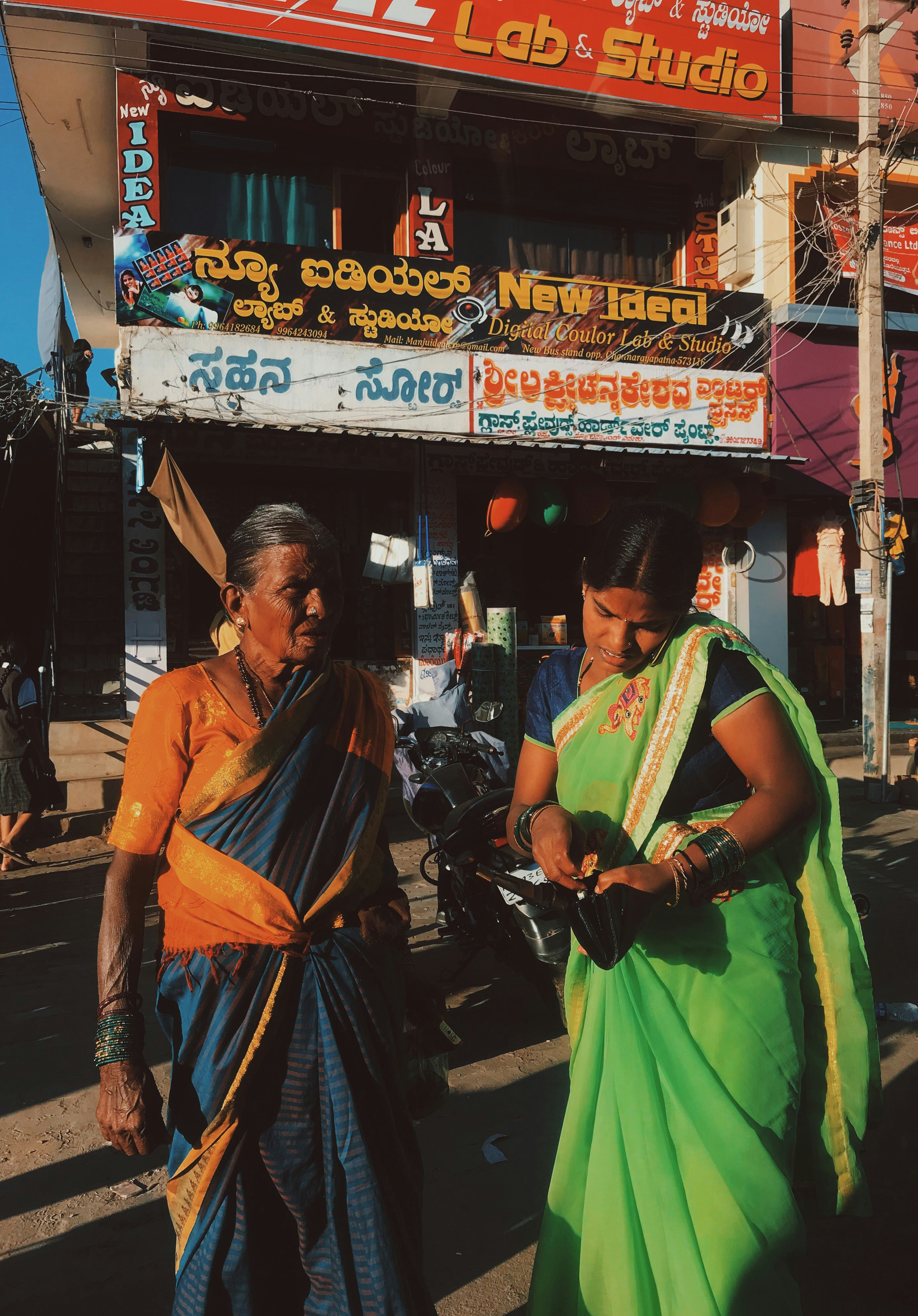 Two women in traditional attire engage in conversation on a bustling street, with colorful shop signs in the background.