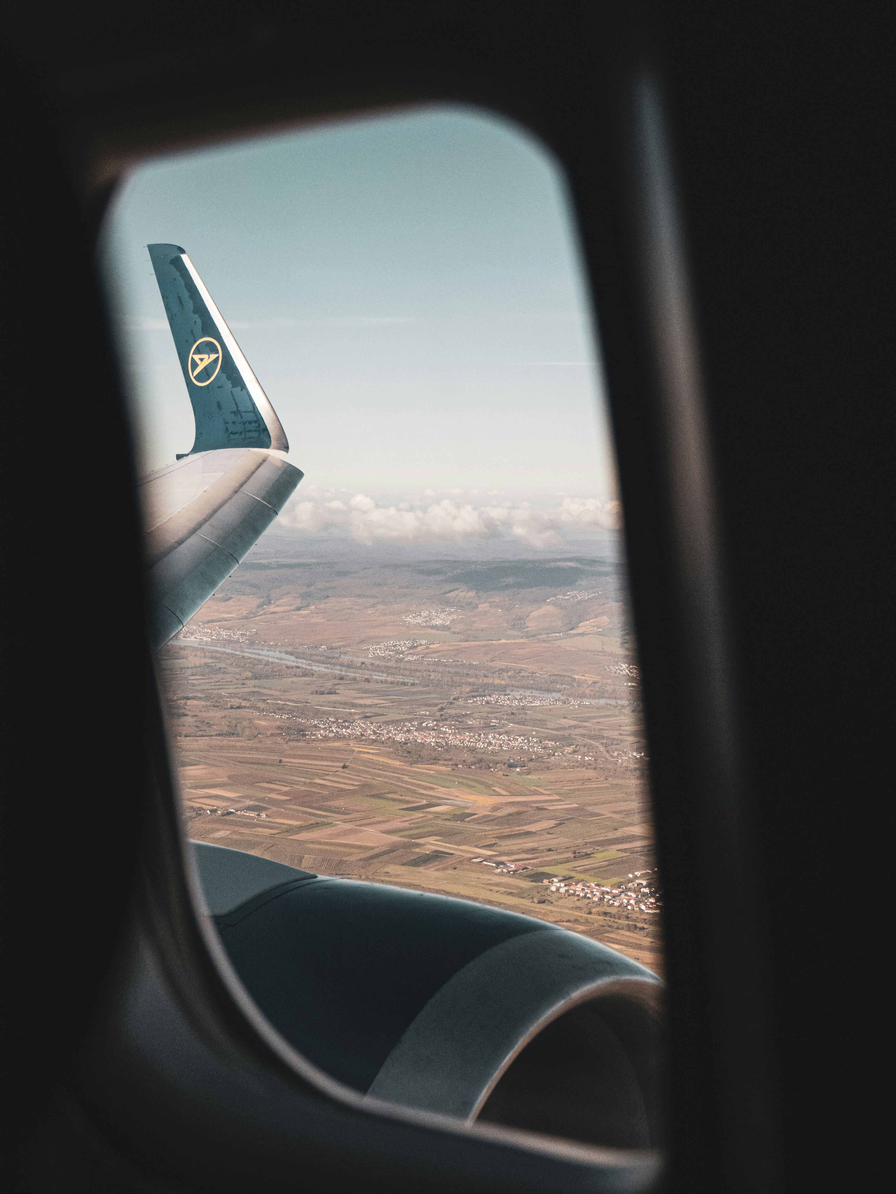 Aircraft wing framed by the airplane window, showcasing a patchwork of fields and distant hills beneath a clear sky.