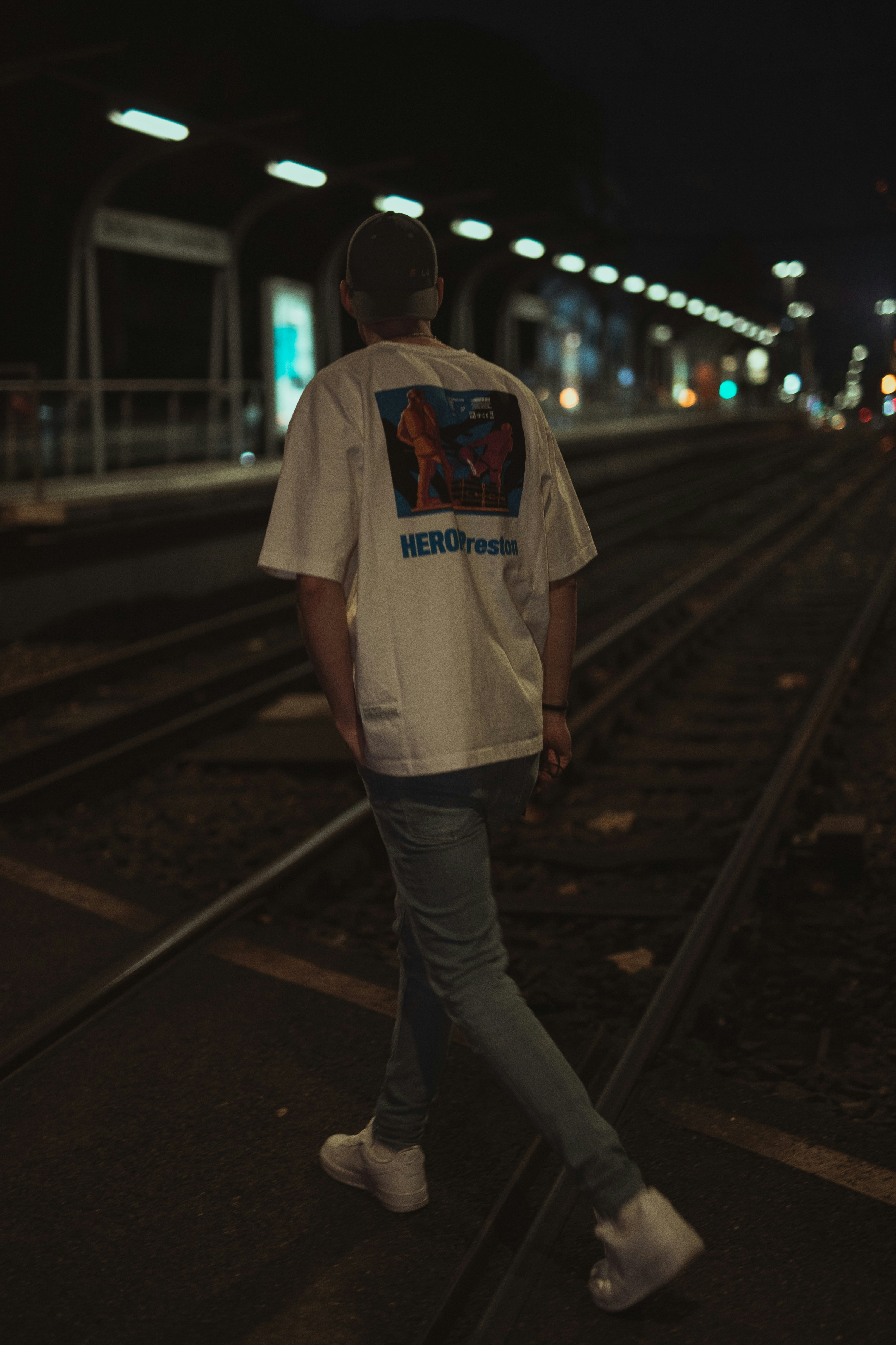 Individual walking along train tracks at night, illuminated by distant lights. The figure wears a graphic tee featuring a vibrant design.