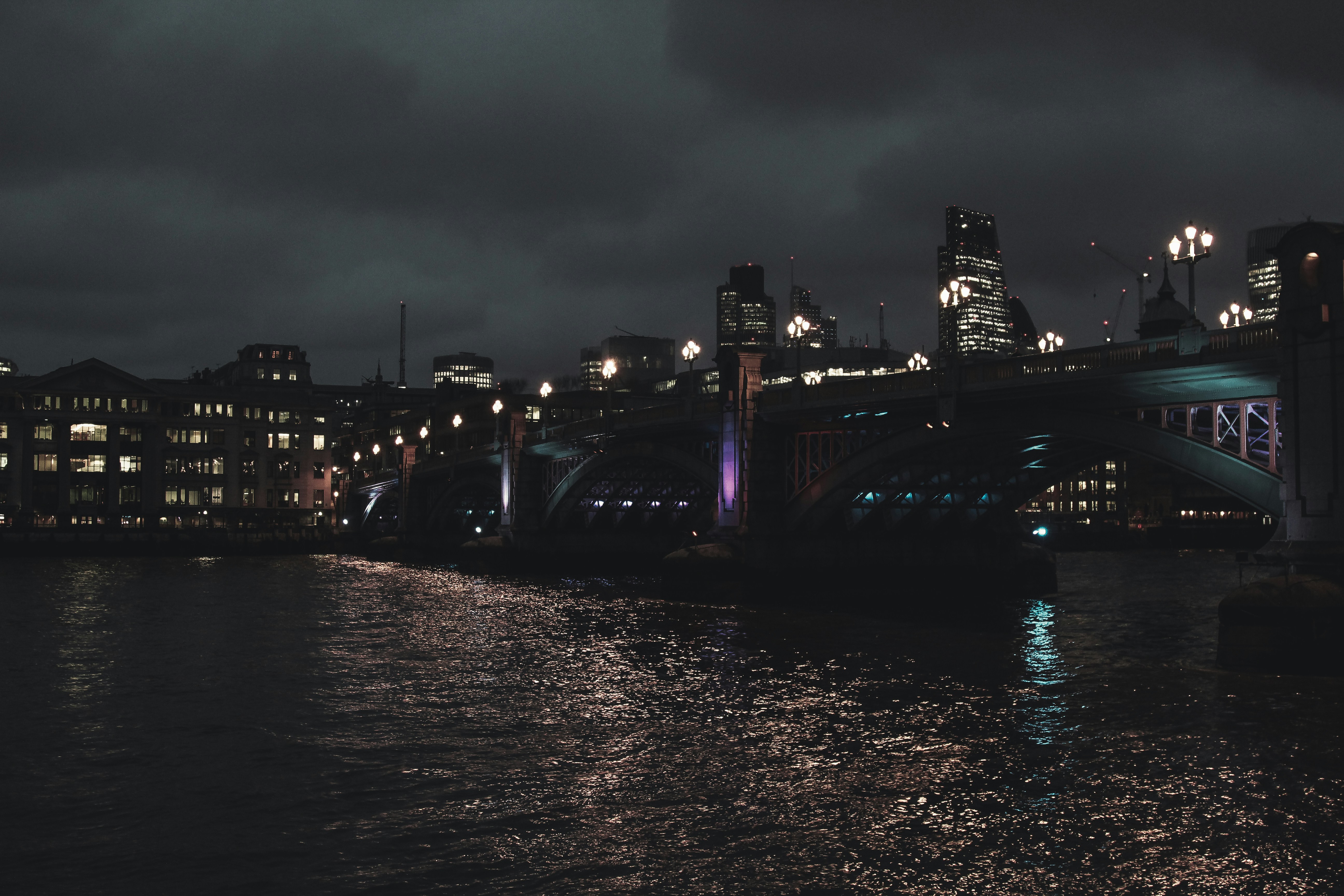 Illuminated bridge spanning the Thames River under a moody sky, with city lights shimmering on the water's surface.