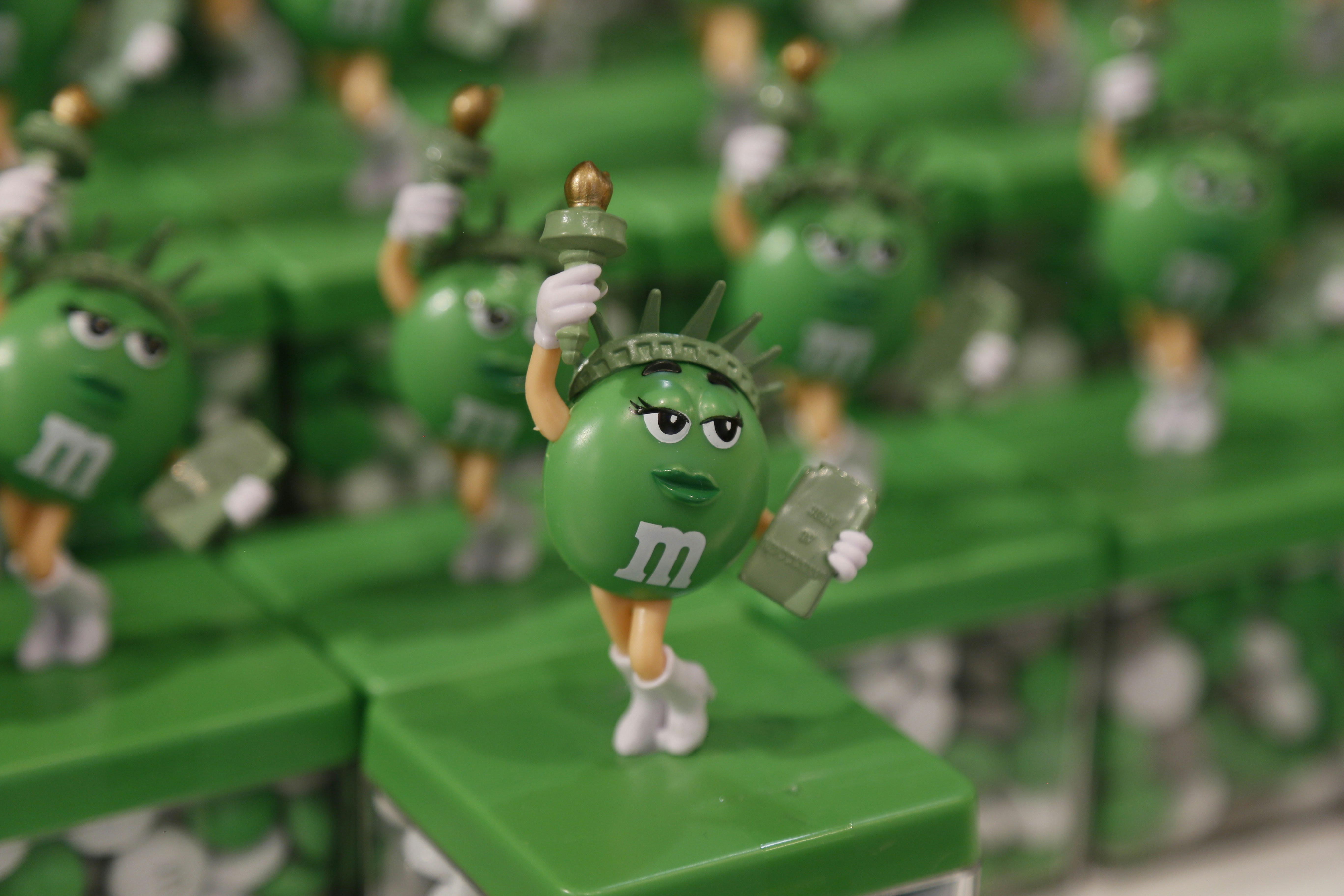 Green M&M's character styled as the Statue of Liberty, holding a torch and tablet, surrounded by a backdrop of similar figures.