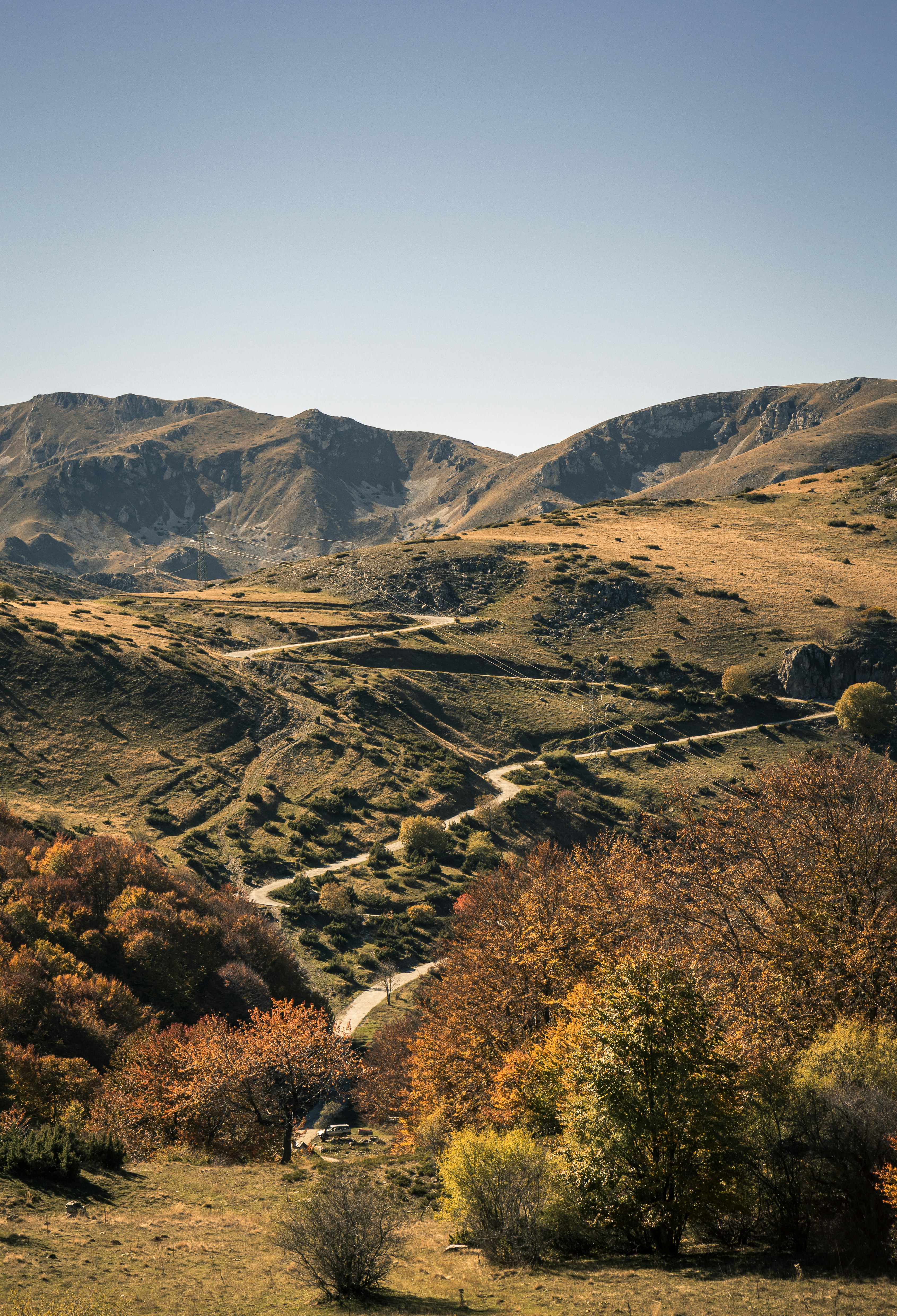 Autumn landscape with a winding dirt road weaving through rolling hills. The foreground shows colorful trees as the distant mountains rise under a clear blue sky.