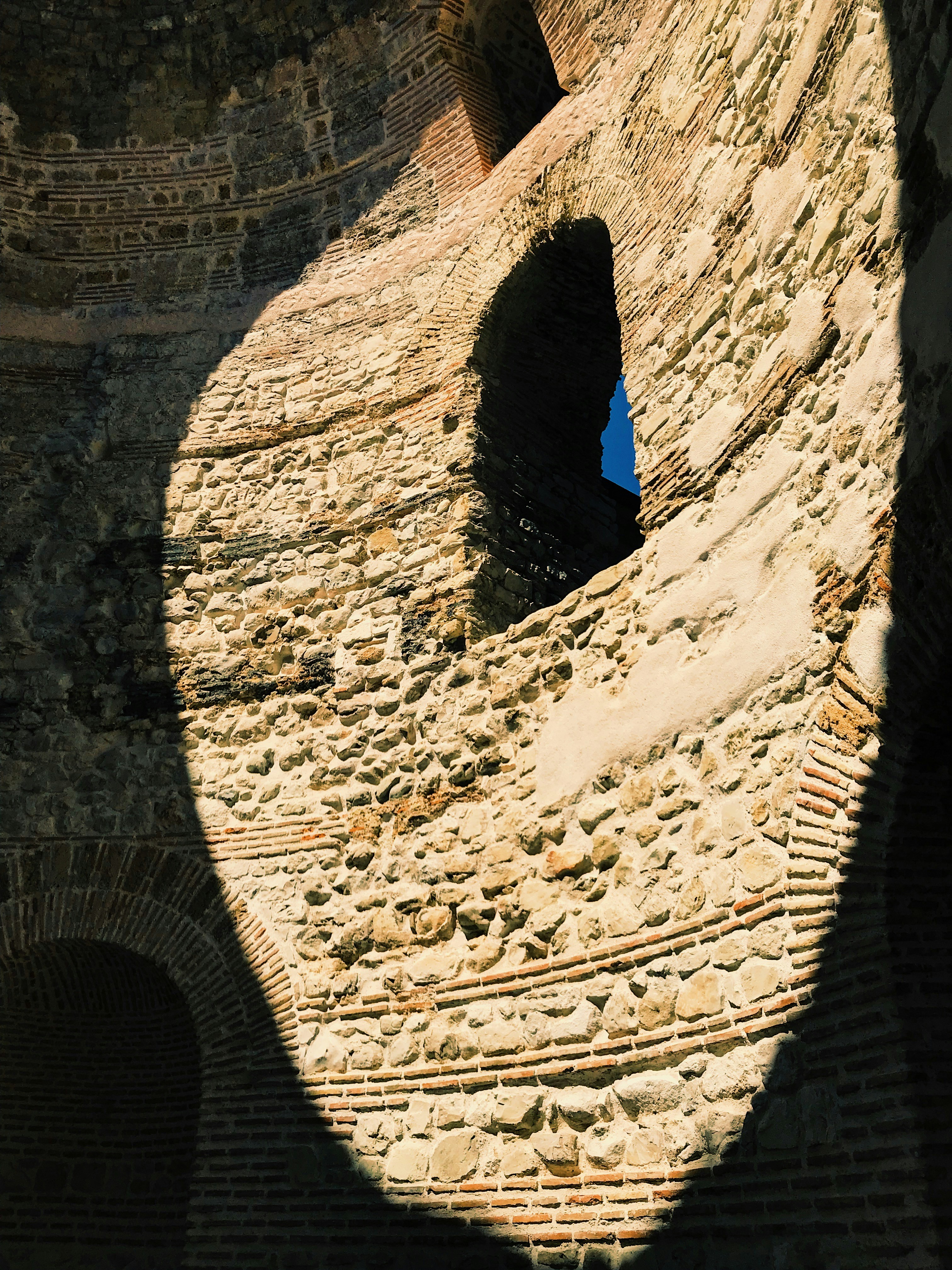 Intricate stonework of a historic structure illuminated by sunlight, revealing a blue sky through an arched opening.