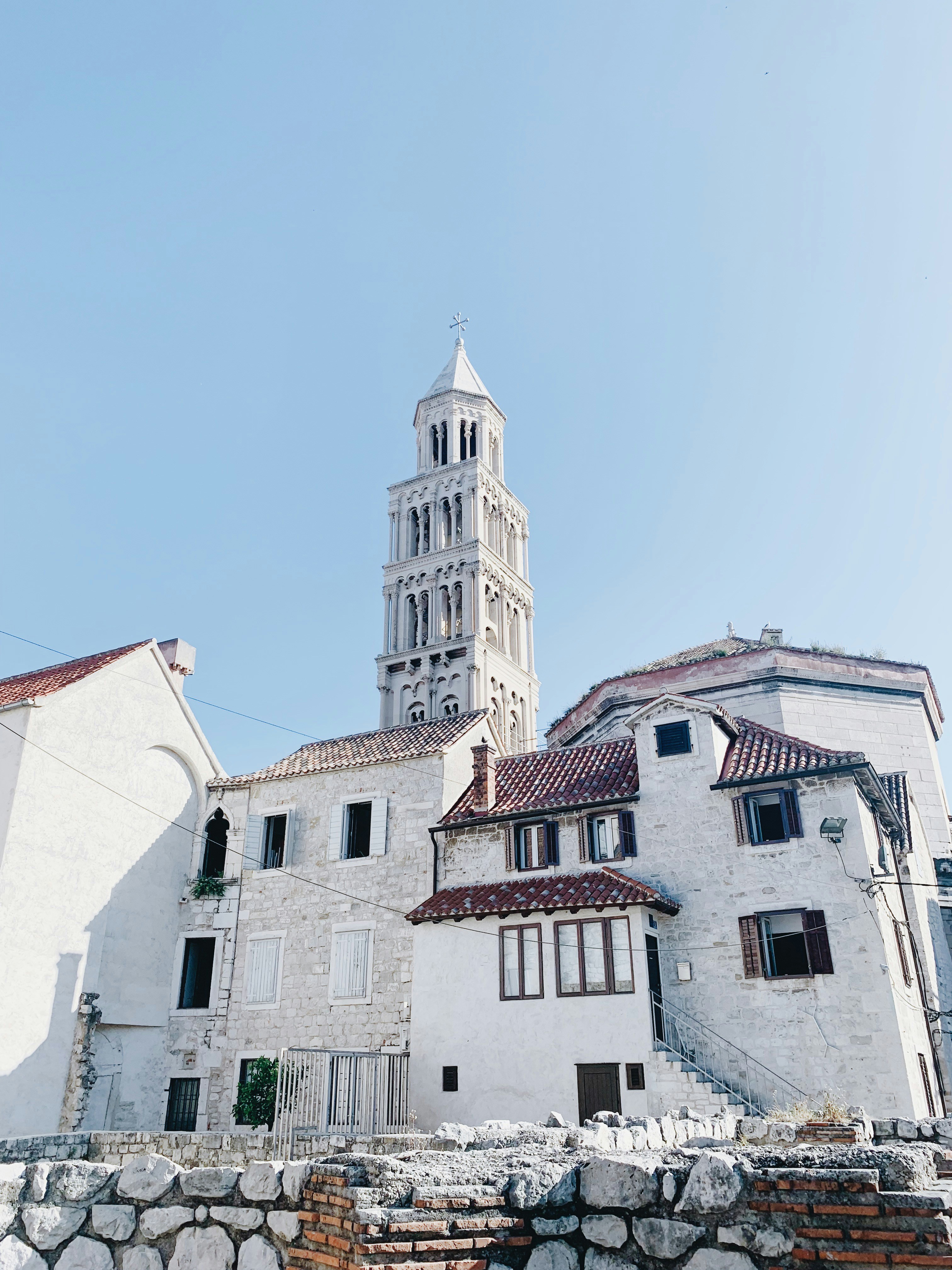 Historic stone buildings with terracotta roofs beneath a clear blue sky, featuring a prominent bell tower. The scene captures the charm of ancient architecture.
