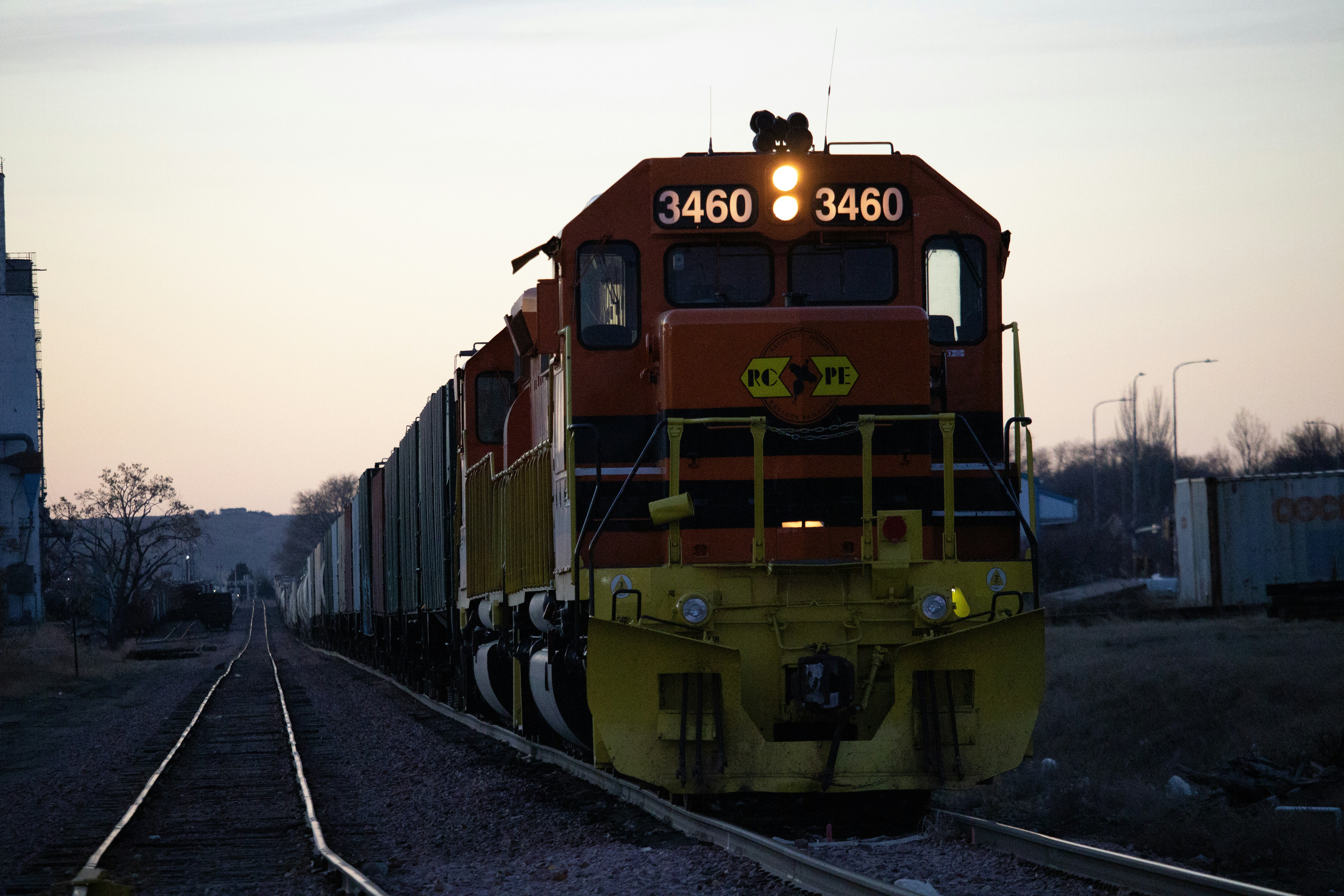 Freight train with bright headlights approaching a station at dusk, showcasing its powerful presence against a fading sky.