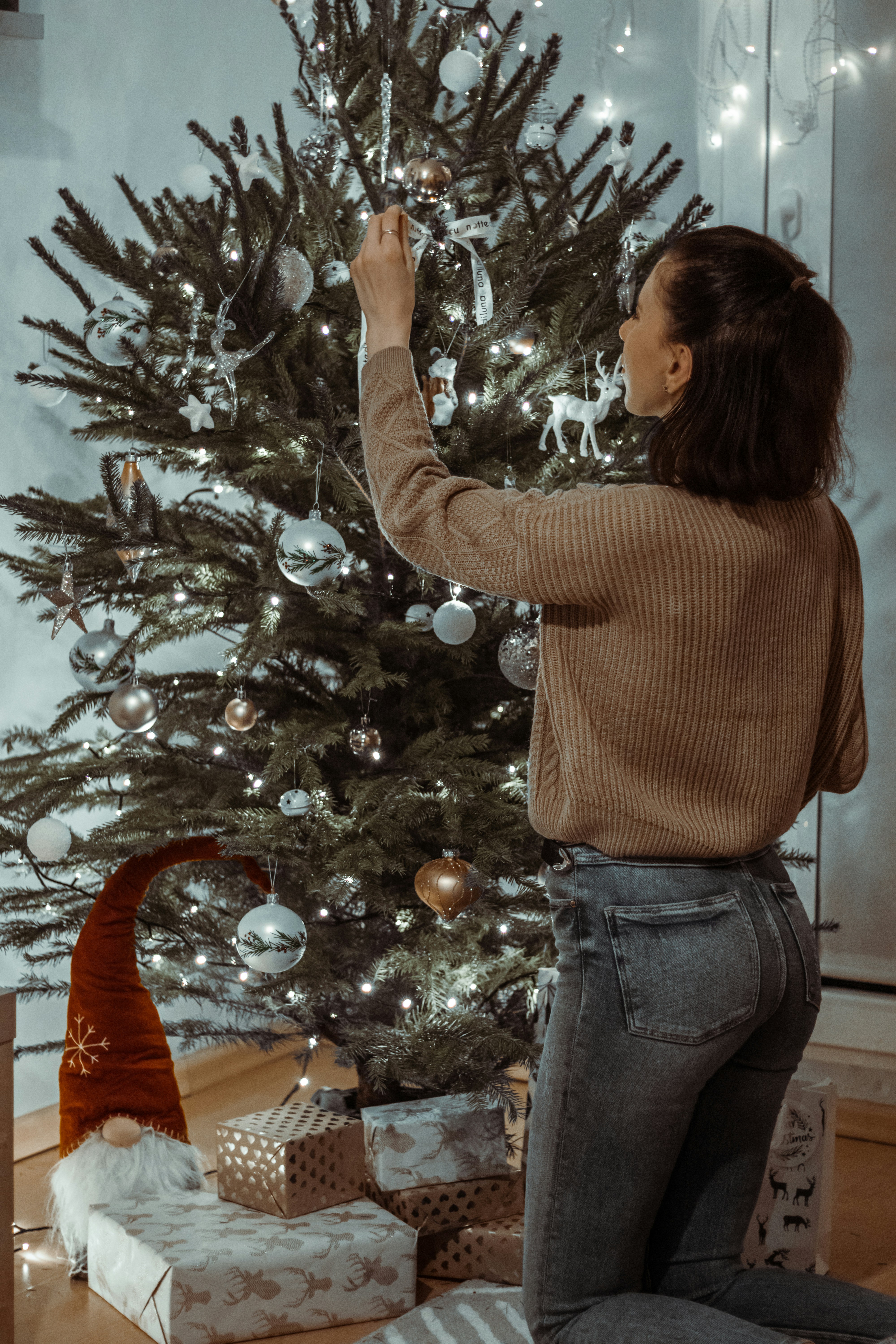 A woman decorates a Christmas tree adorned with ornaments and lights, while festive gifts are neatly arranged beneath it.