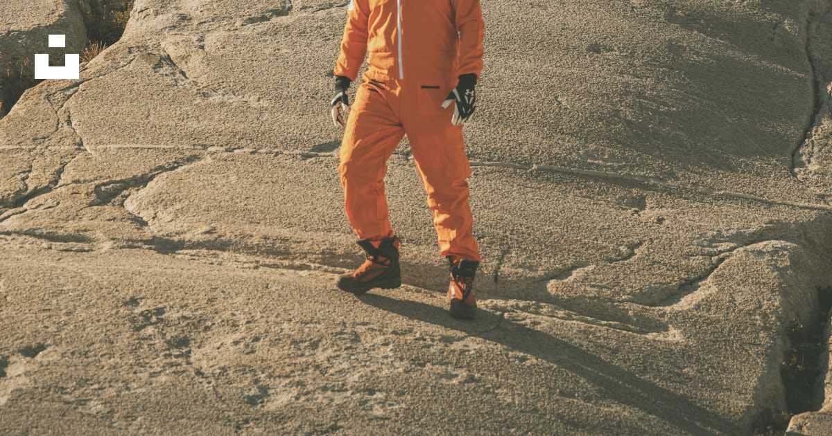 Man In Orange Jacket And Pants Walking On Brown Sand During Daytime