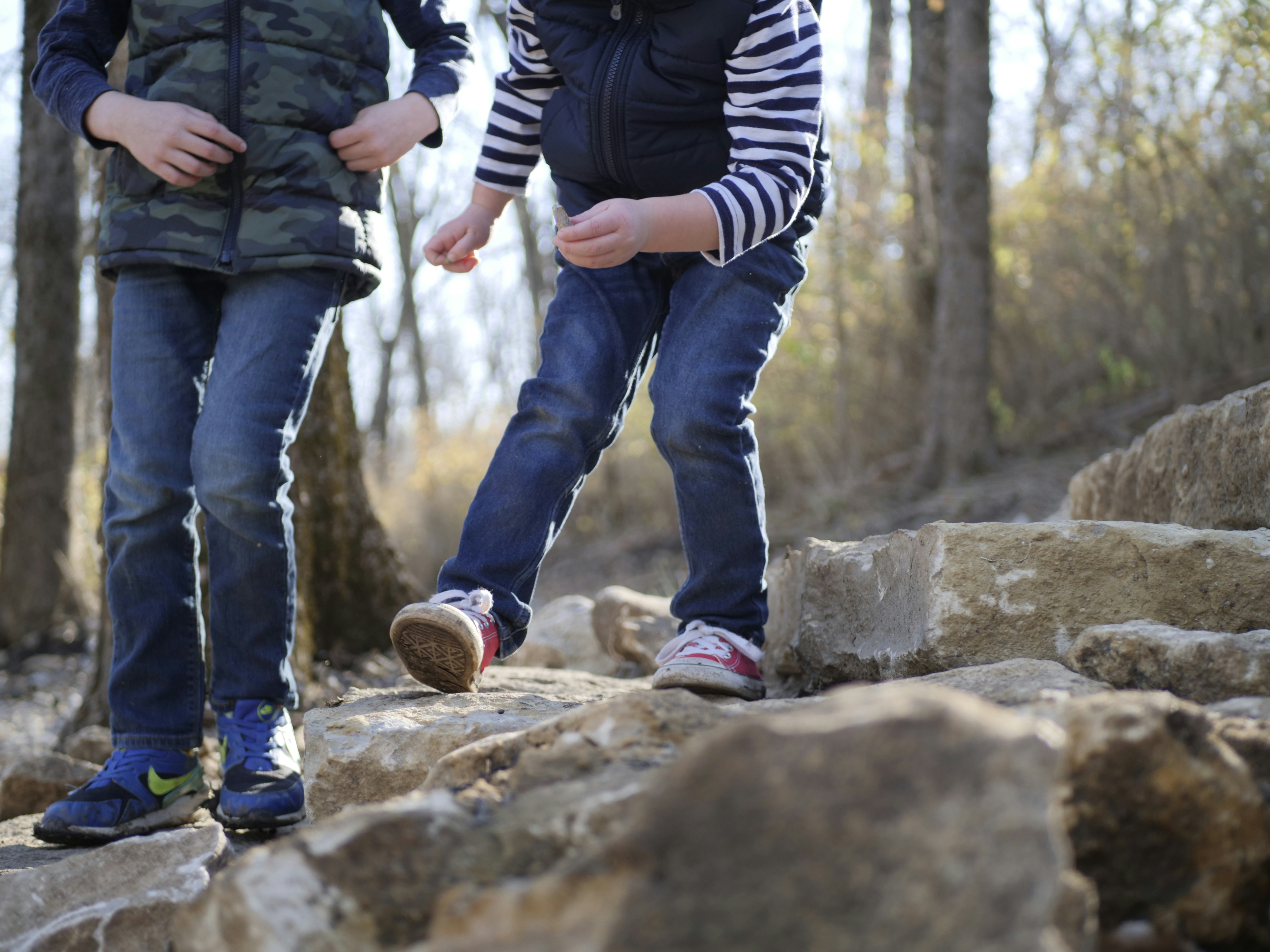 Two kids stride across sunlit rocks on a woodland trail