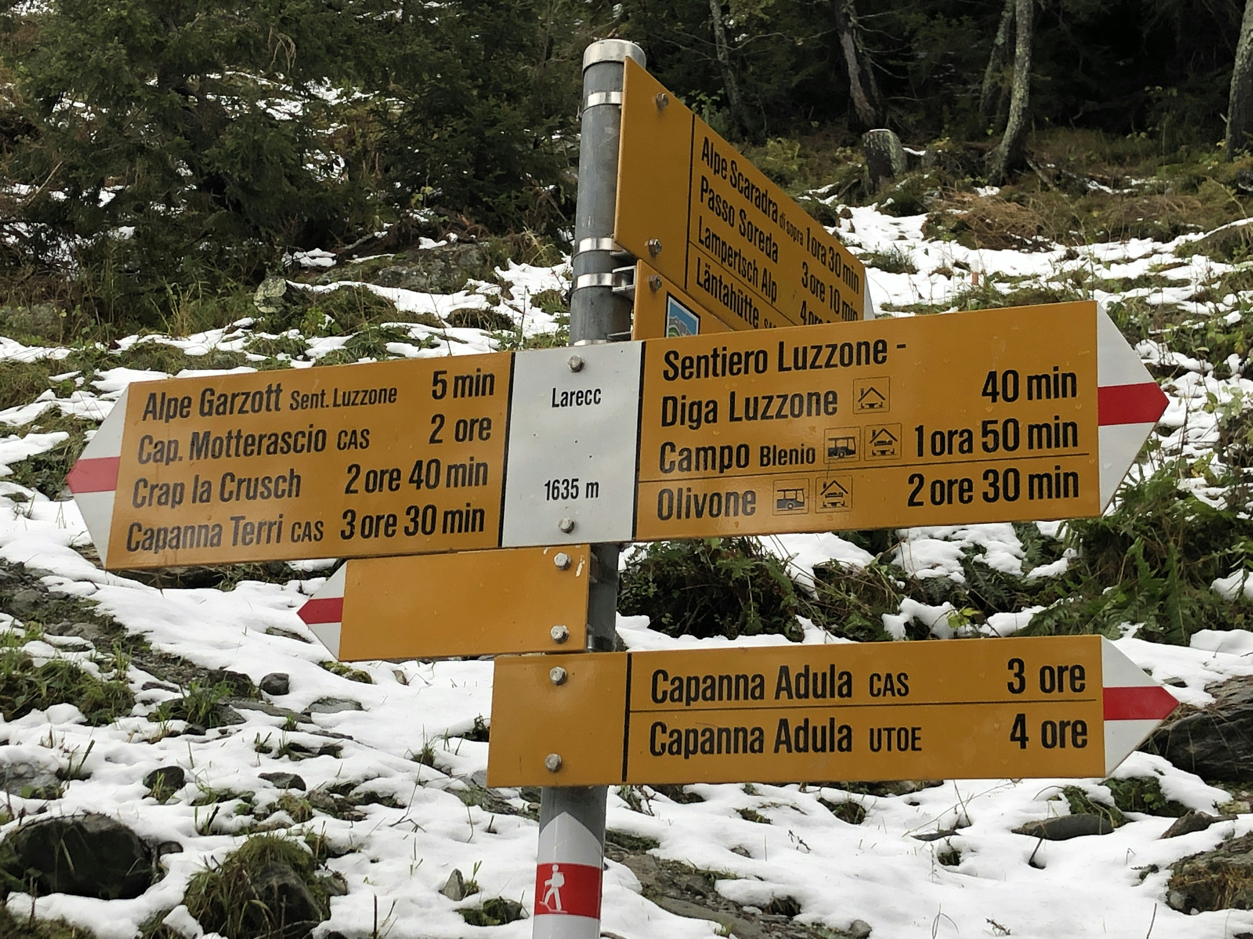 Signpost displaying various hiking trail directions and estimated times in a snowy alpine setting.