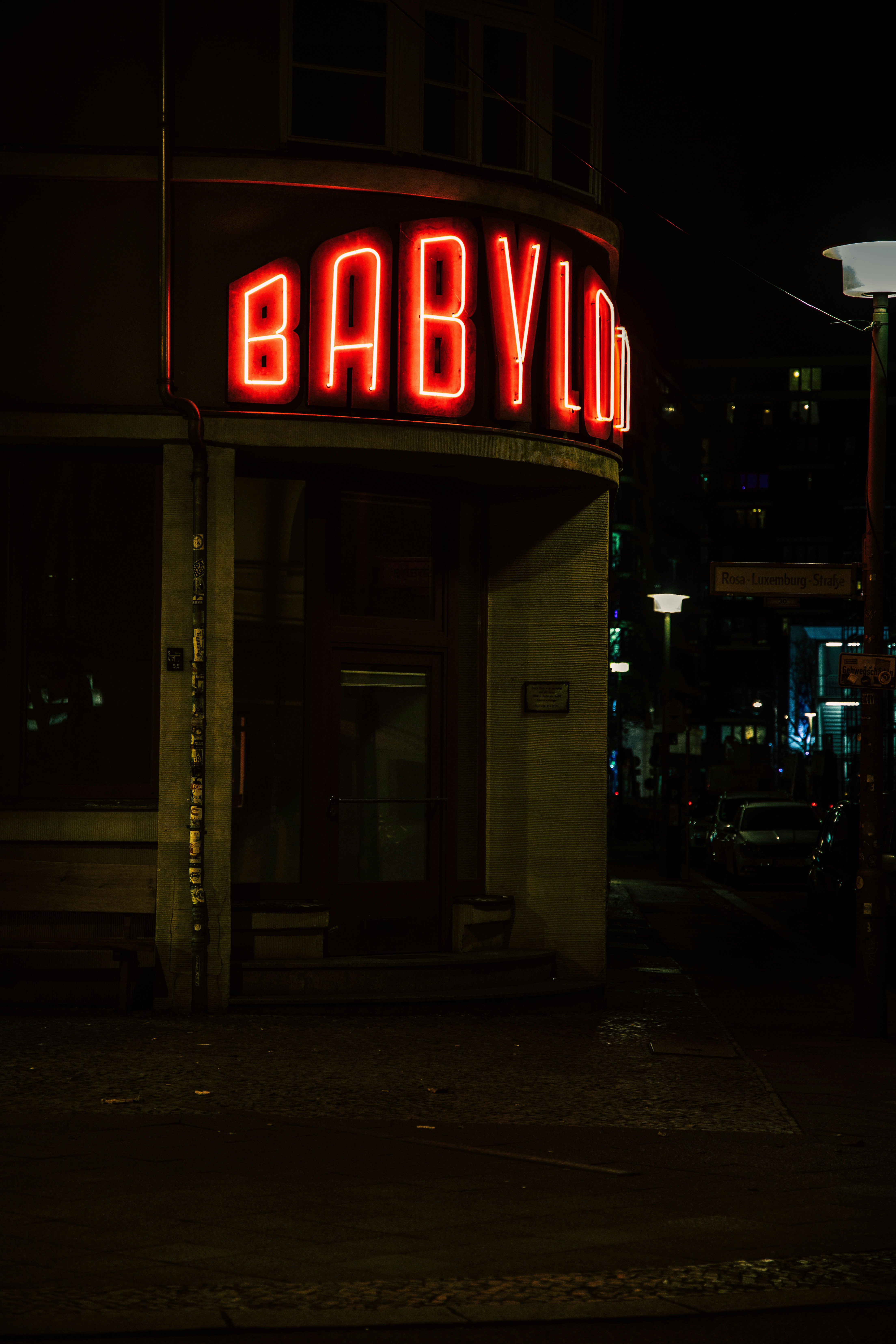 Illuminated red neon sign reading 'BABYLON' on a dimly lit street corner at night.