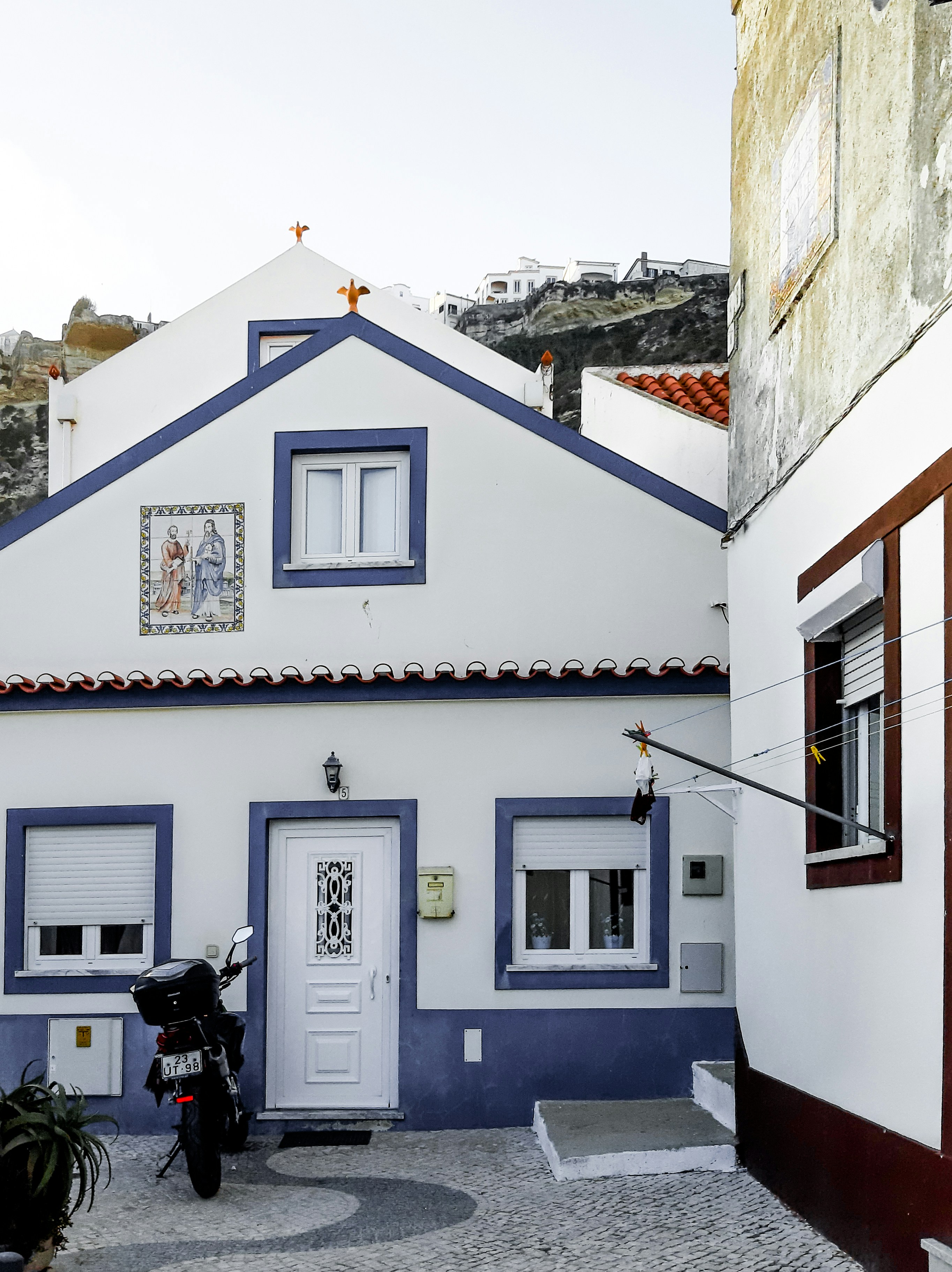 A quaint white house adorned with blue accents and a decorative mural, nestled in a picturesque alleyway. The scene captures the essence of coastal architecture.