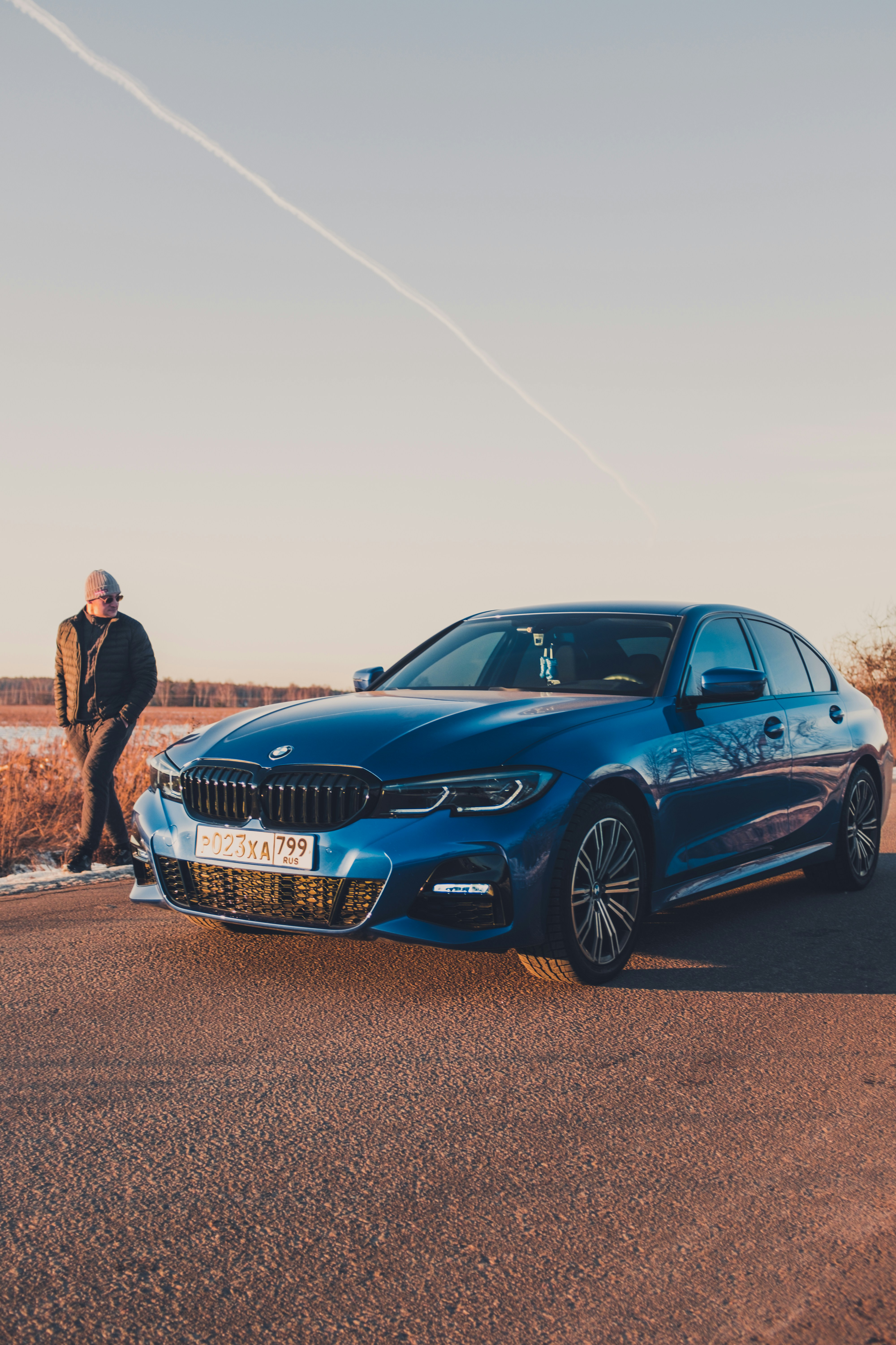 A sleek blue BMW parked by a serene landscape, with a figure walking in the background under a clear sky.
