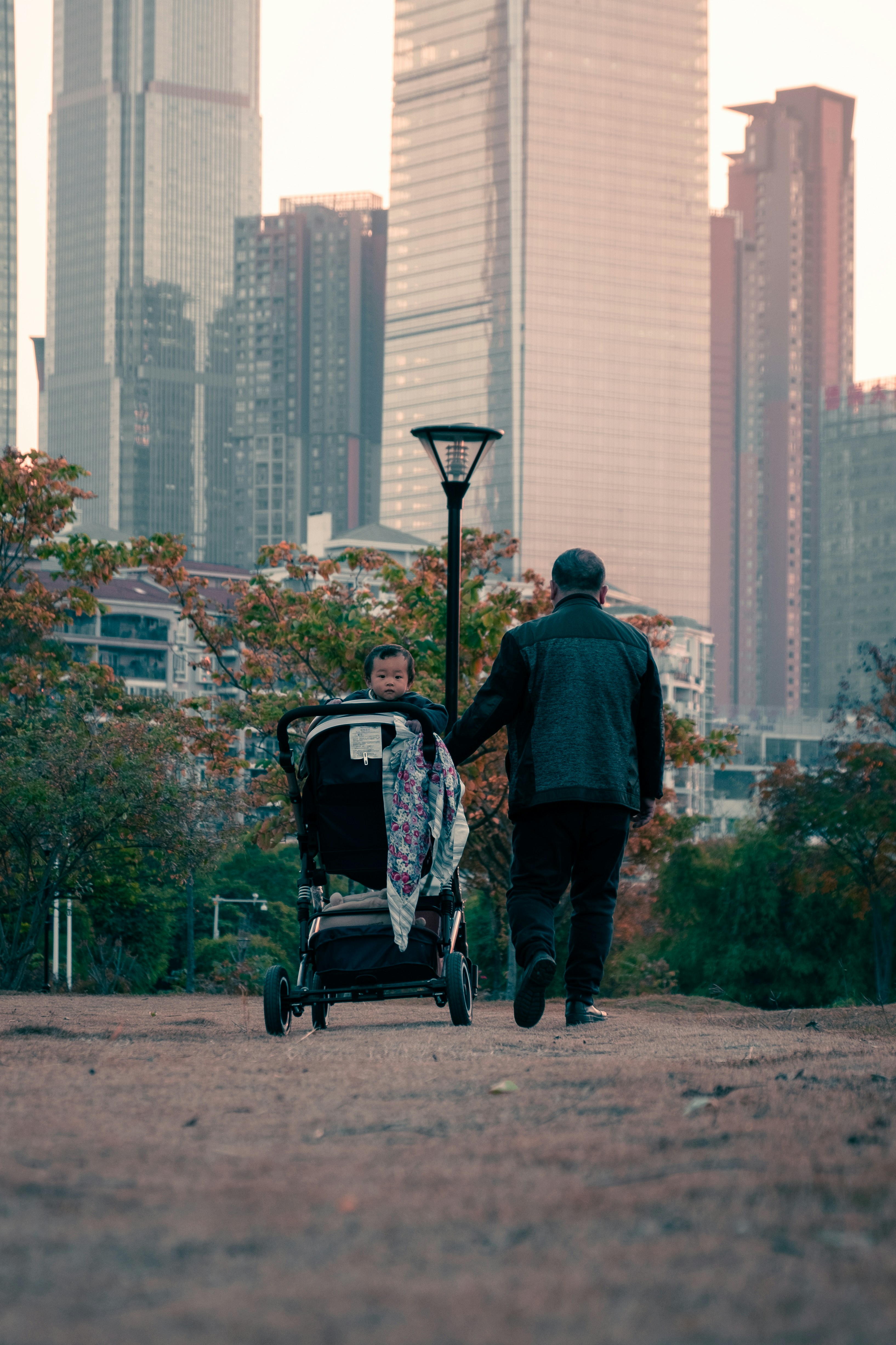 An elderly man pushes a child in a stroller along a path, framed by towering city skyscrapers and autumn foliage. The scene captures a moment of connection amid urban development.
