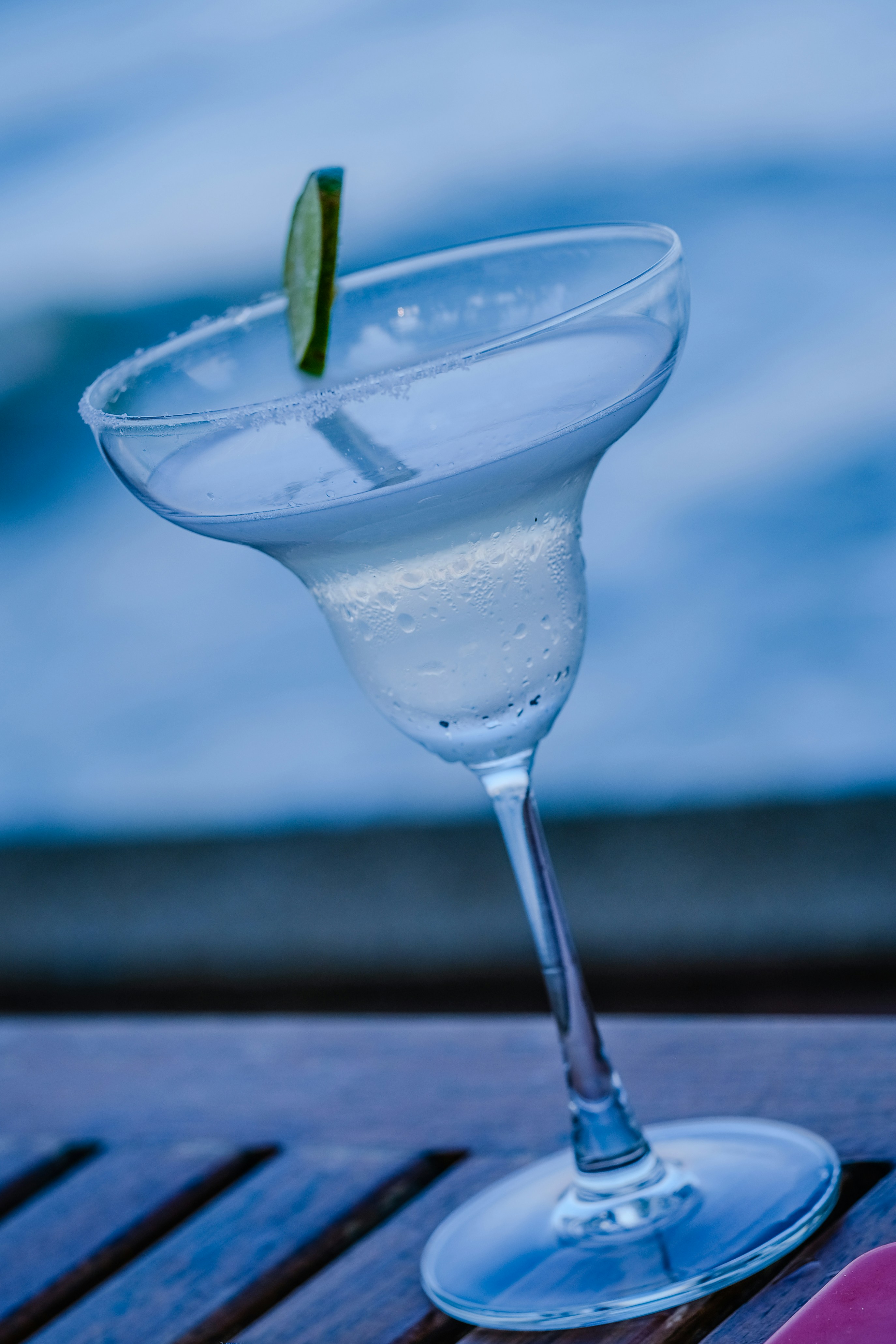 A refreshing margarita garnished with a lime slice, resting on a wooden table with a blurred ocean backdrop.