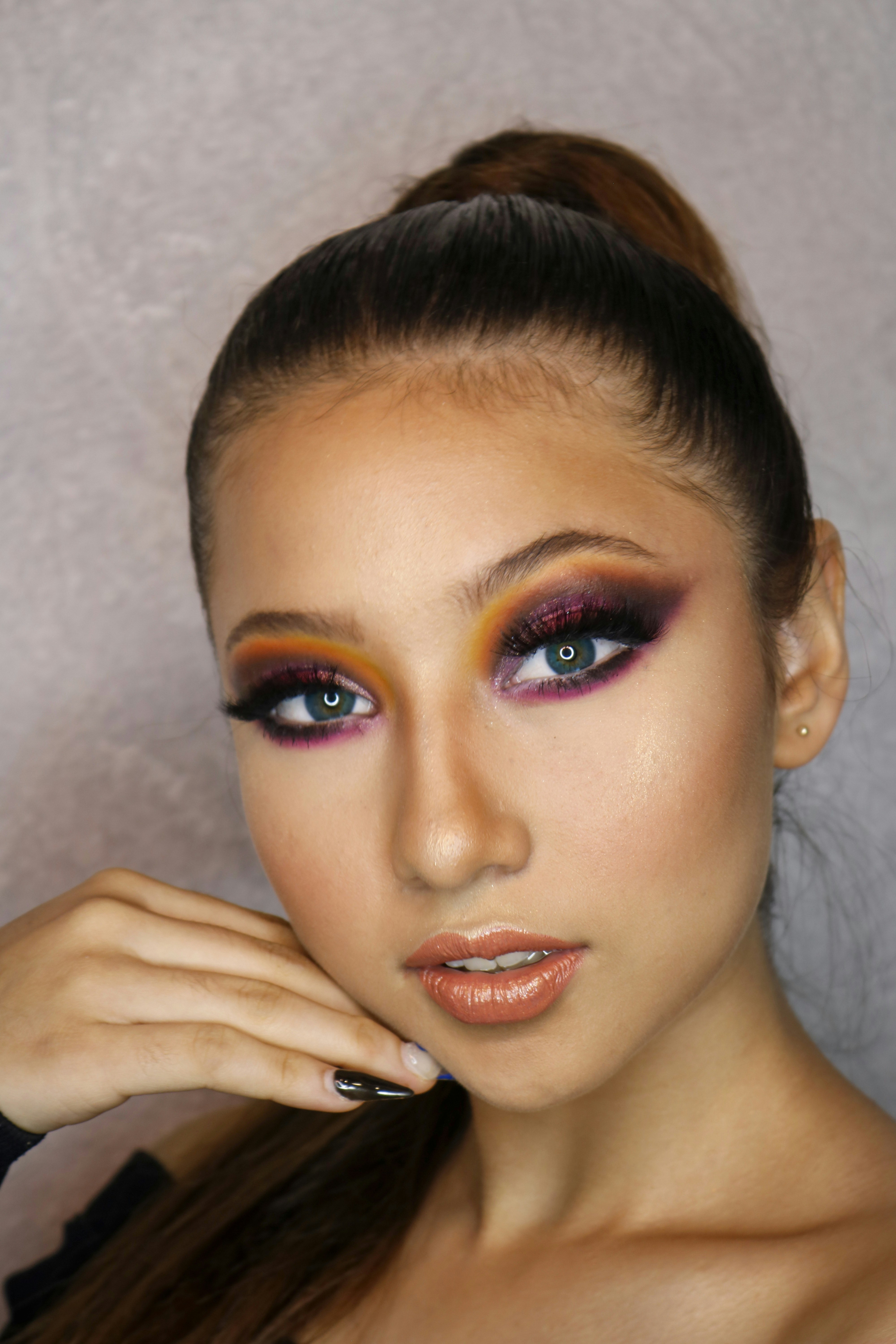 Close-up portrait featuring a model with striking makeup showcasing bold colors and intricate details. The model's pose adds a touch of sophistication.