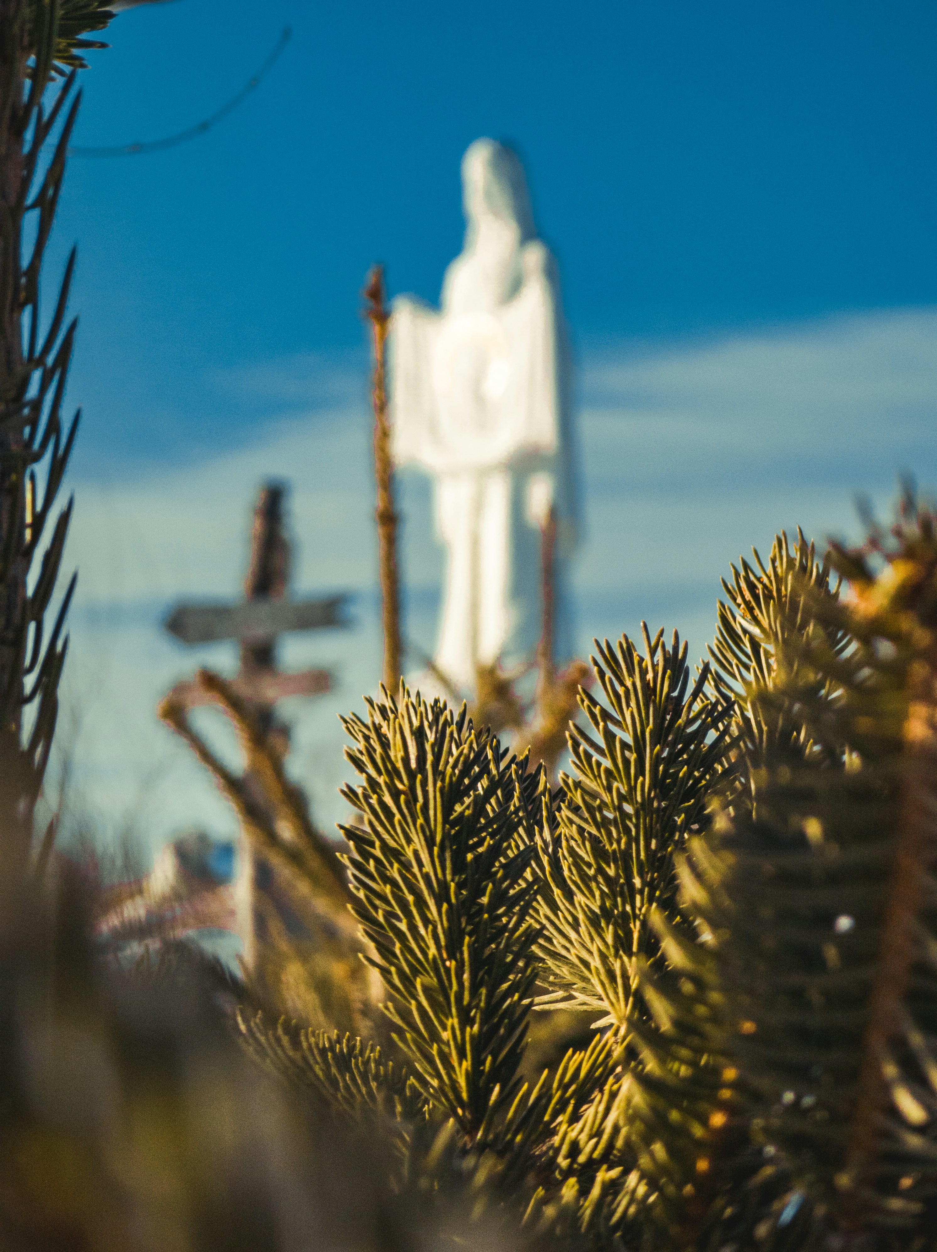 A white statue of a figure stands tall against a blue sky, framed by vibrant greenery in the foreground.