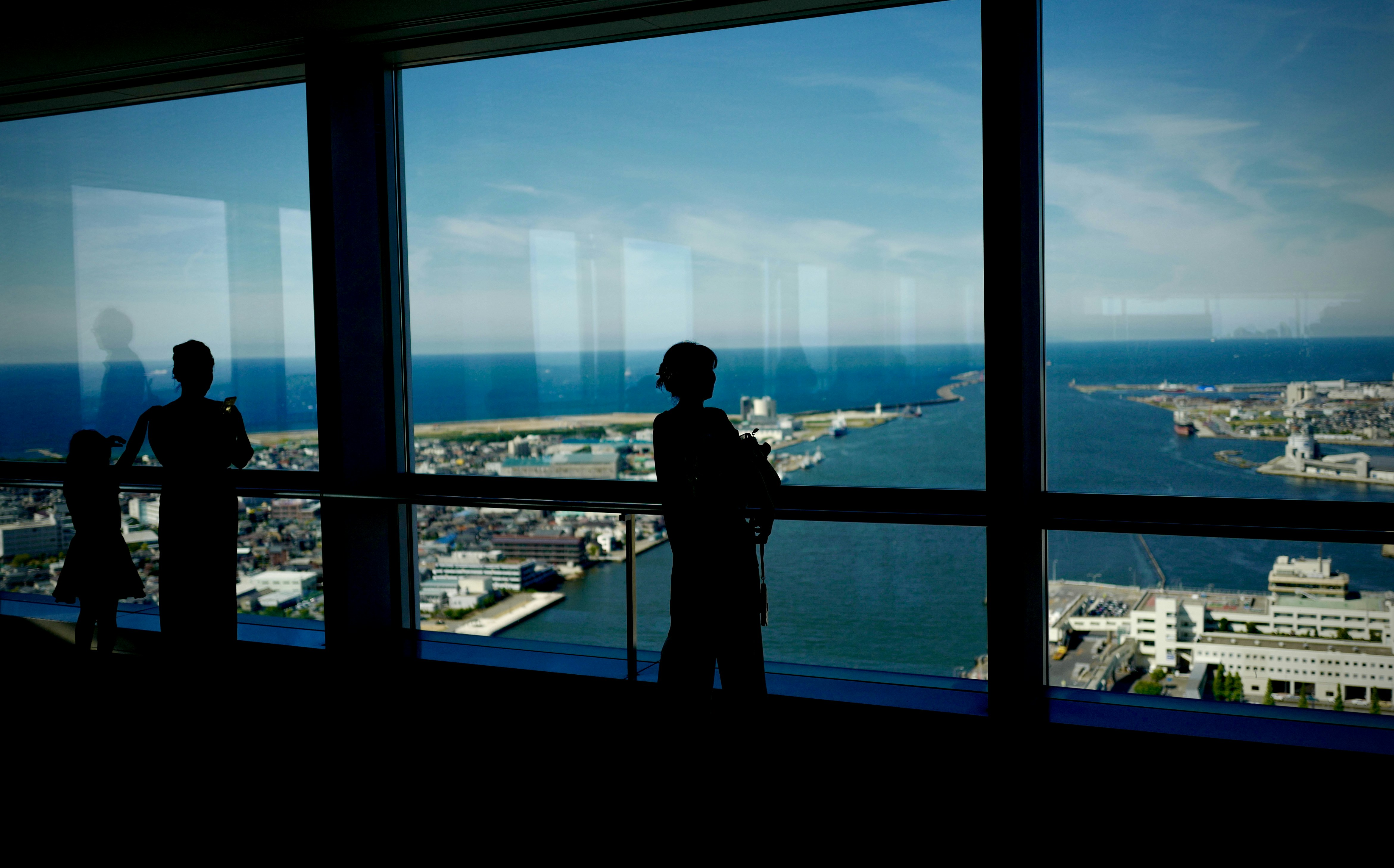Silhouetted figures stand before large windows overlooking a coastal cityscape and expansive waterway.