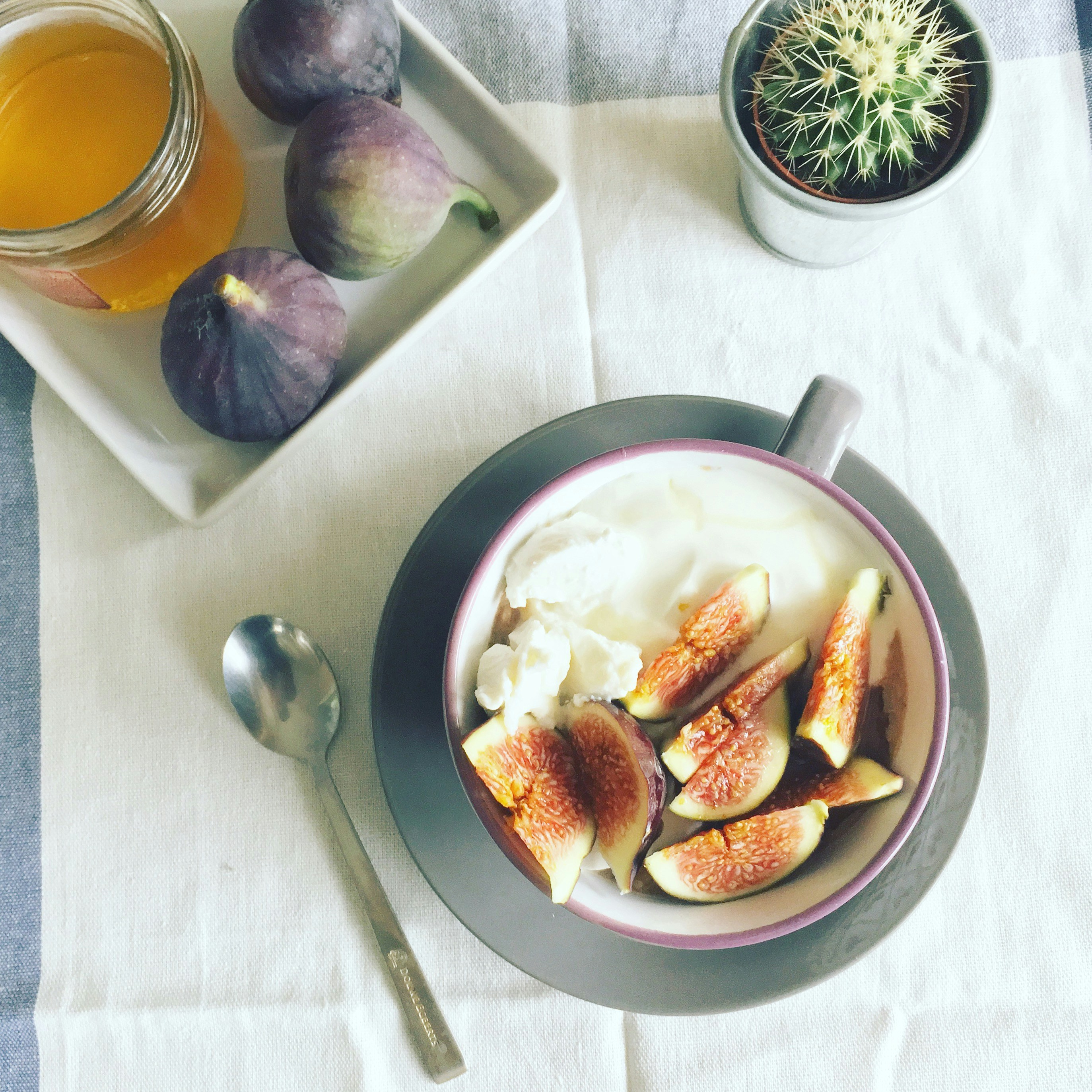 Bowl of fresh figs and yogurt accompanied by a jar of honey and a plate of figs, set on a striped tablecloth with a small cactus in a pot.