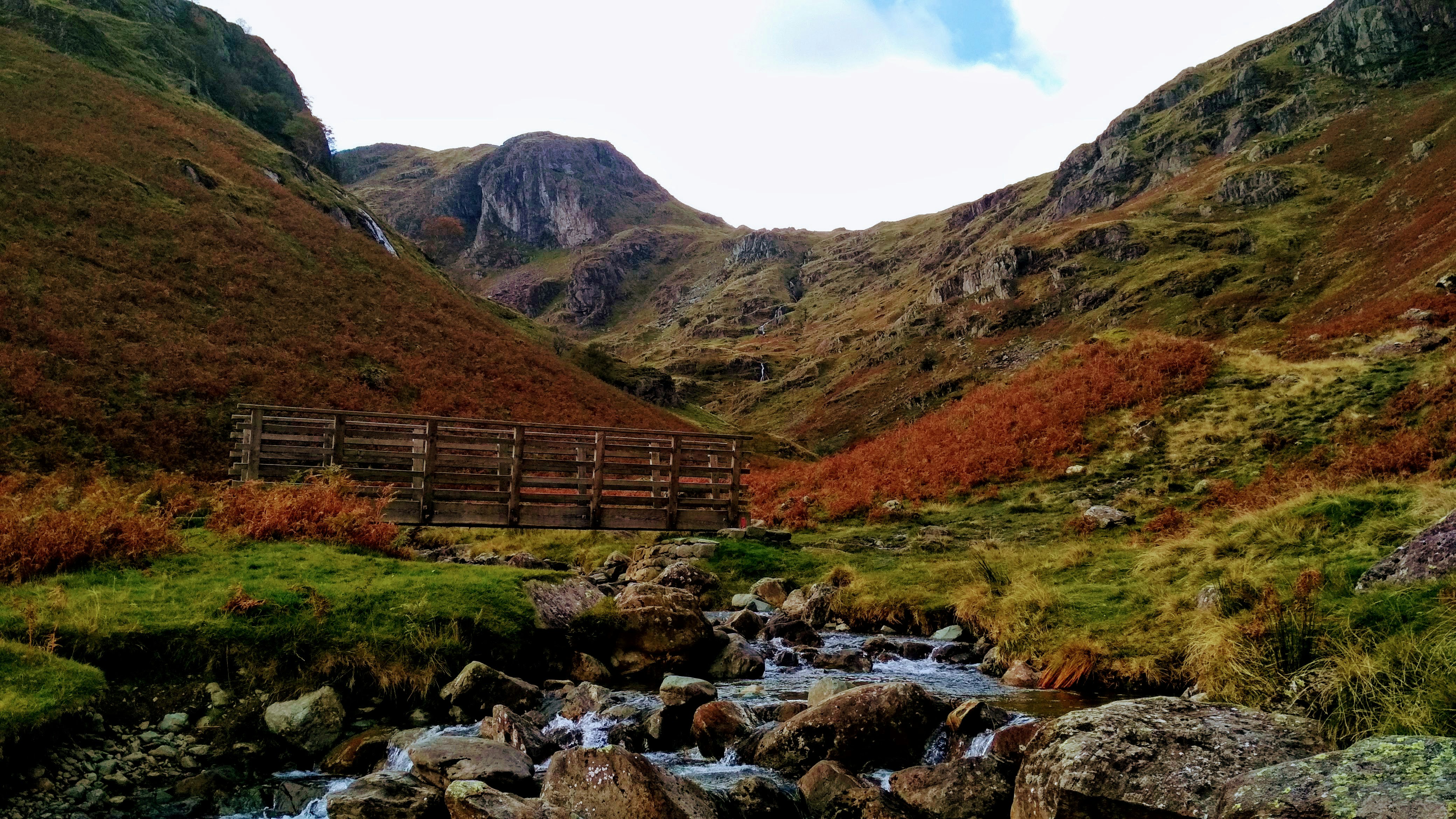 Wooden fence bordering a serene stream in a vibrant autumn landscape, framed by rolling hills and rocky terrain.