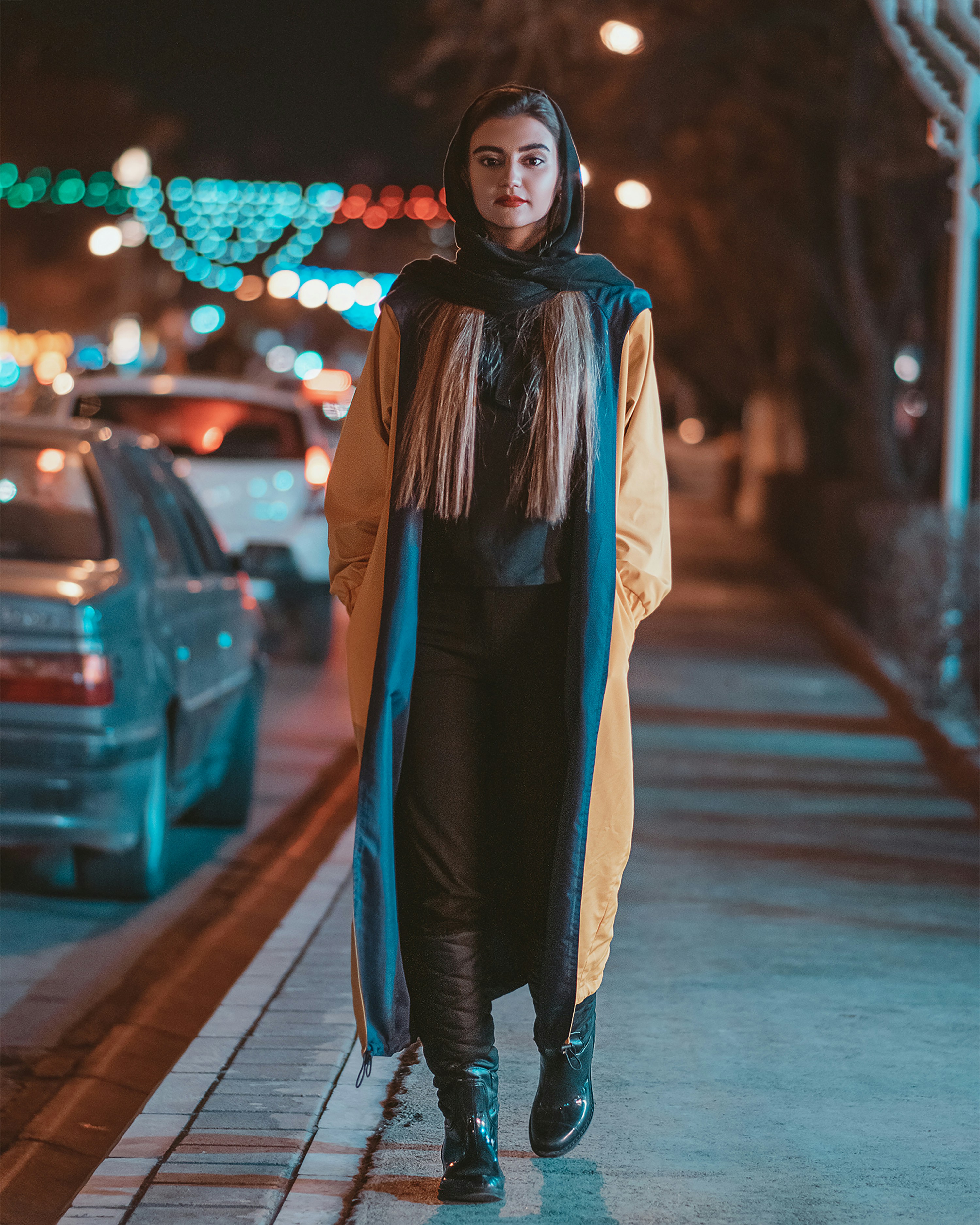 A woman in a vibrant, long coat walks confidently along a city street adorned with colorful lights. The scene captures the essence of urban life at night.