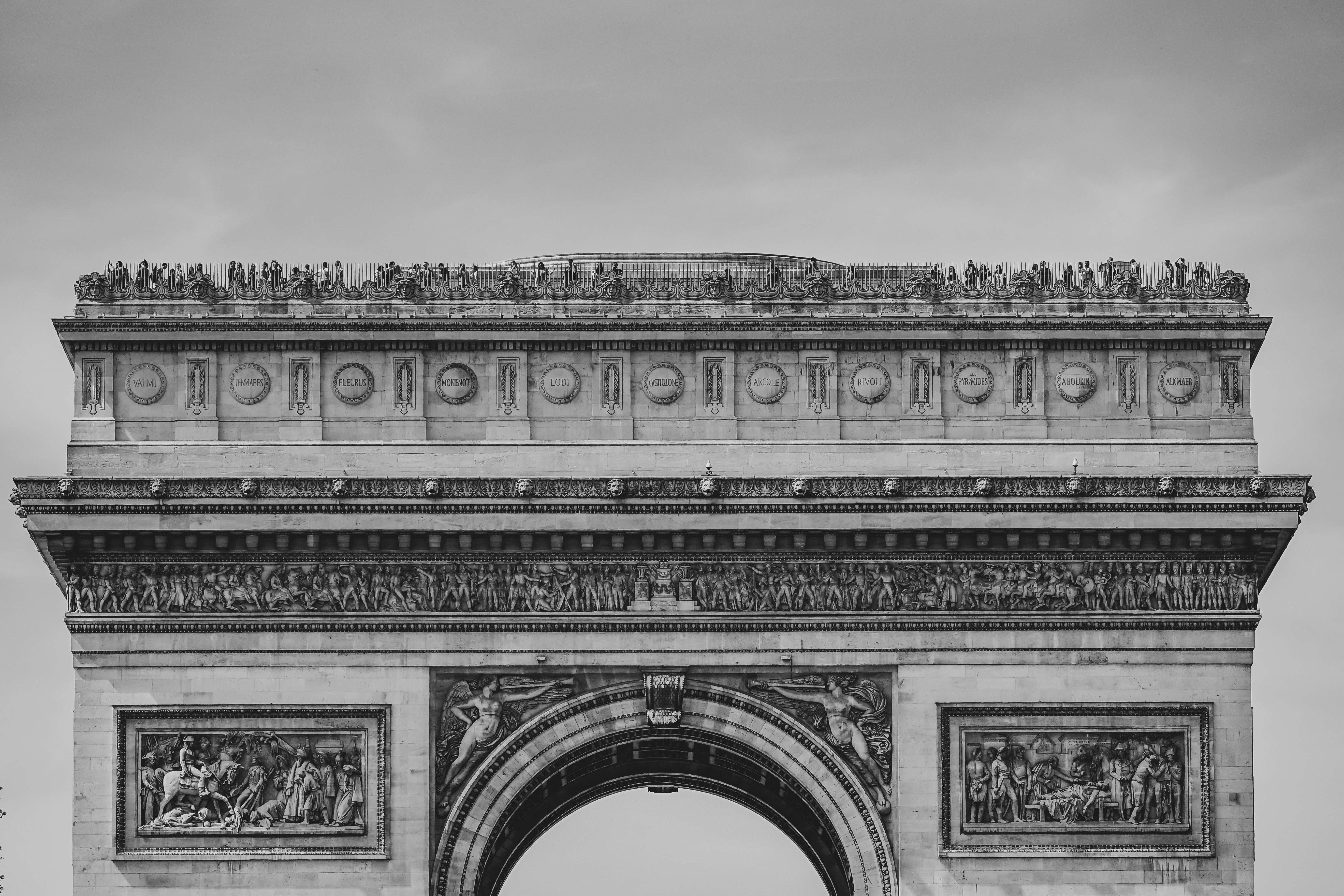 Black-and-white view of the Arc de Triomphe's upper facade, with a line of visitors along the parapet. Ornate relief panels and the central arch anchor the composition.