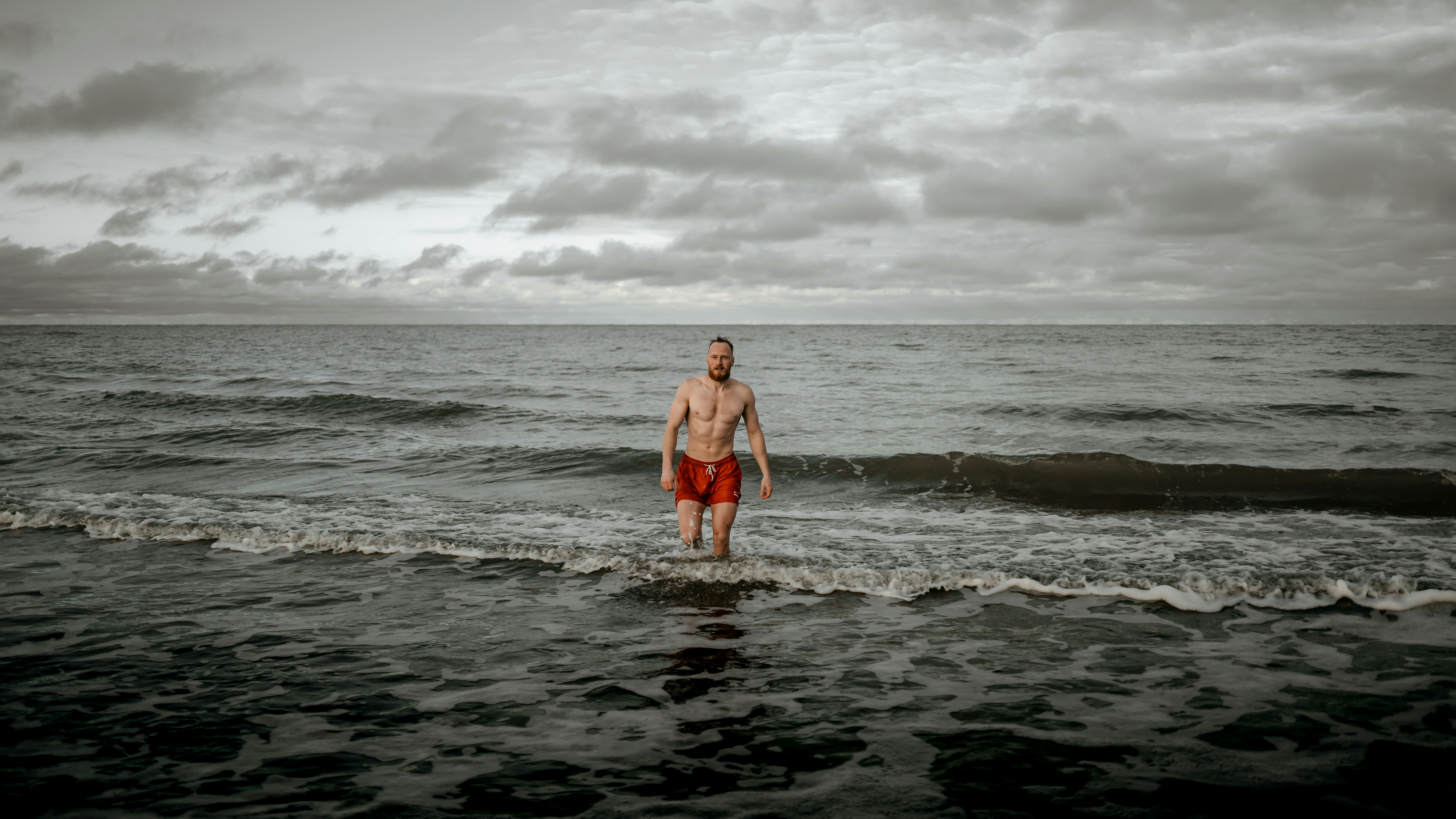 A man in red shorts walks through the waves, with a dramatic sky and ocean backdrop. The scene captures the raw beauty of nature and human resilience.