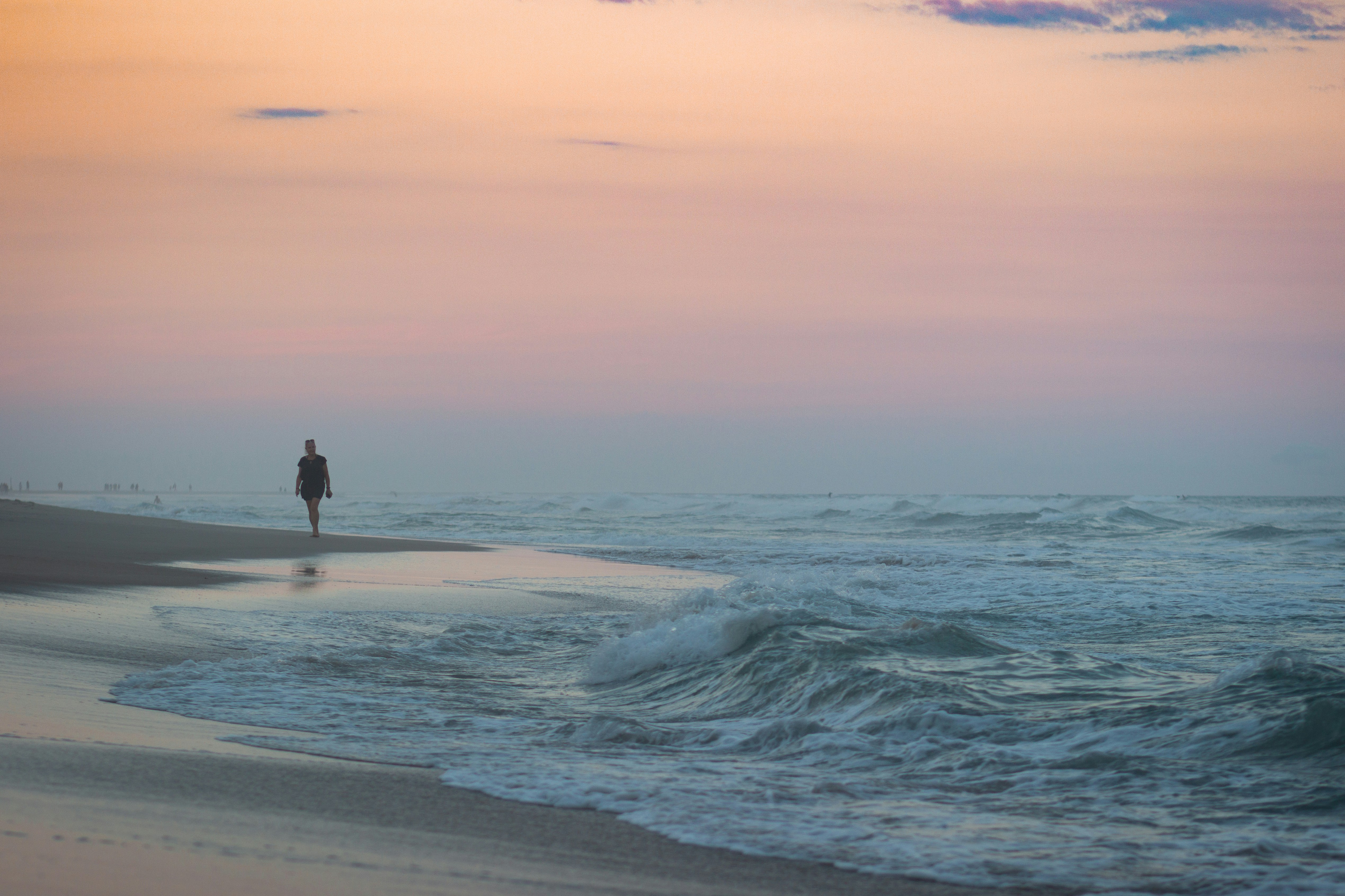 Person walking along a tranquil beach with gentle waves under a pastel sunset sky.