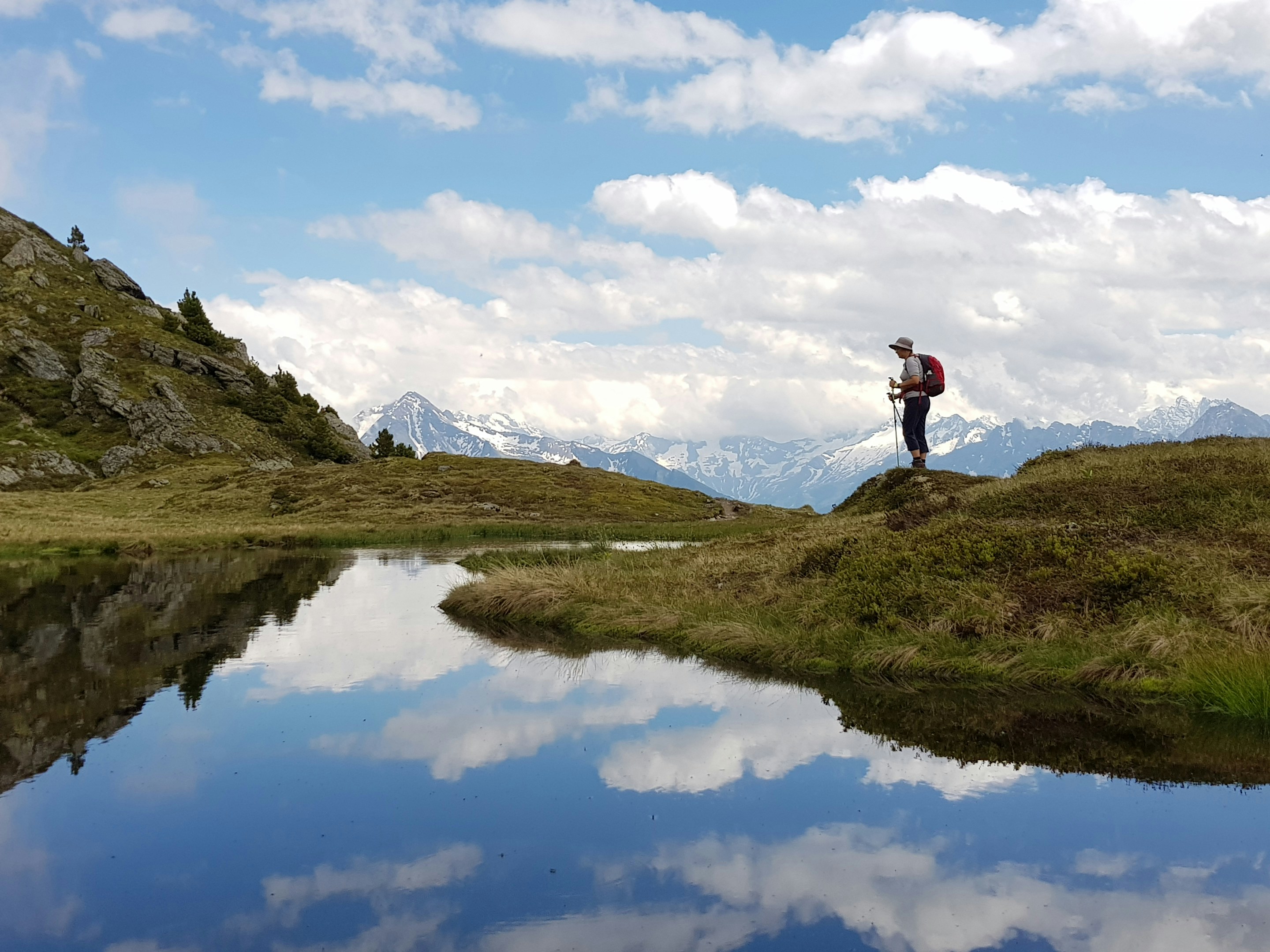 Frau mit rotem Rucksack und Hut an einem glasklaren Bergsee in den Tiroler Alpen. Berglandschaft, Schneeberge und Wolken spiegeln sich im einsamen Teich.
Woman with a red backpack and hat at a crystal-clear mountain lake in the Tyrolean Alps. Mountain landscape, snow peaks and clouds are reflected in the pond. | man in red shirt and black pants standing on green grass field near lake during daytime
