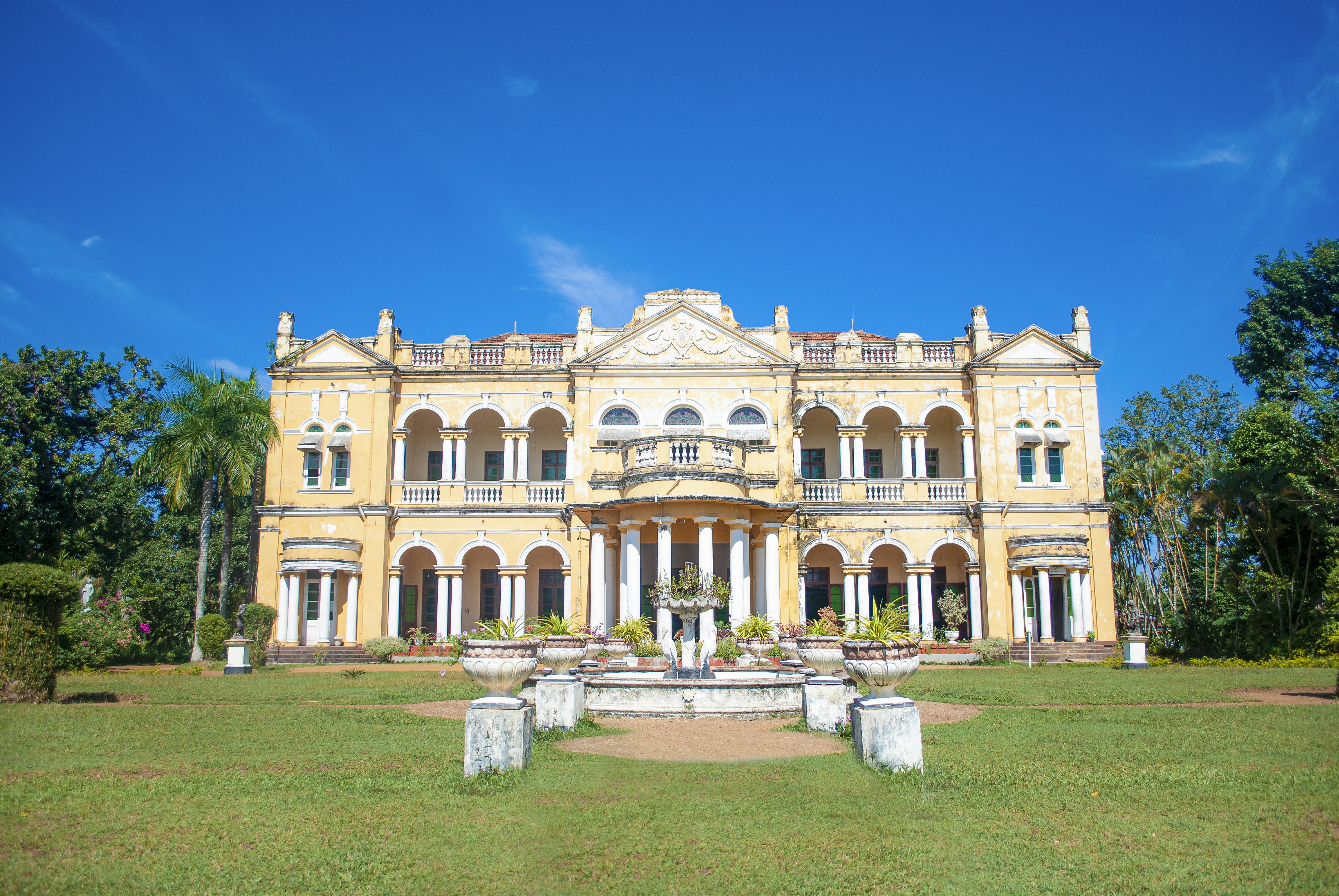 Historic colonial mansion with ornate architecture, surrounded by lush greenery and a clear blue sky.