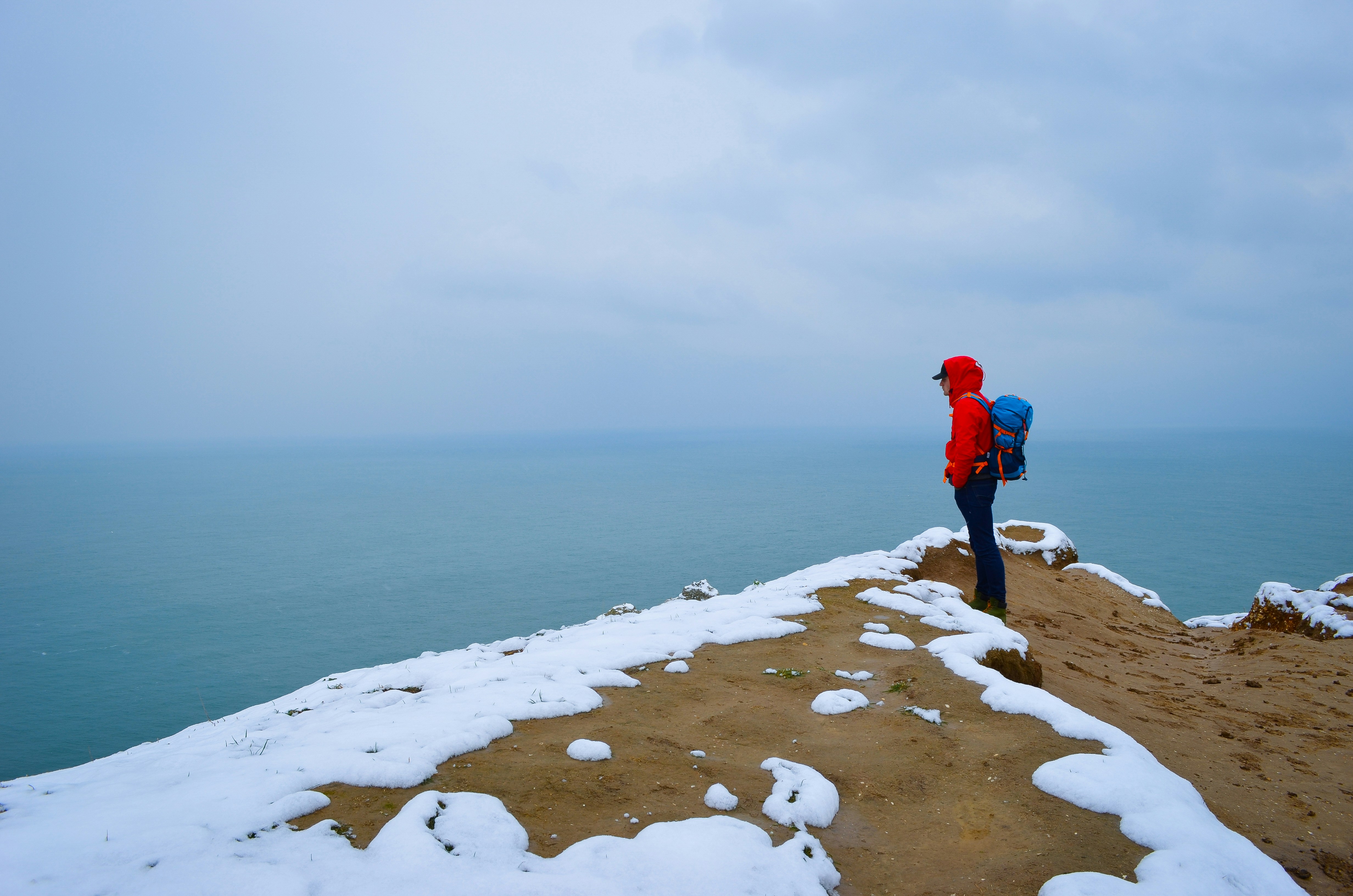 Hiker in a red jacket stands on a snow-covered cliff, gazing out over a calm sea under a cloudy sky.