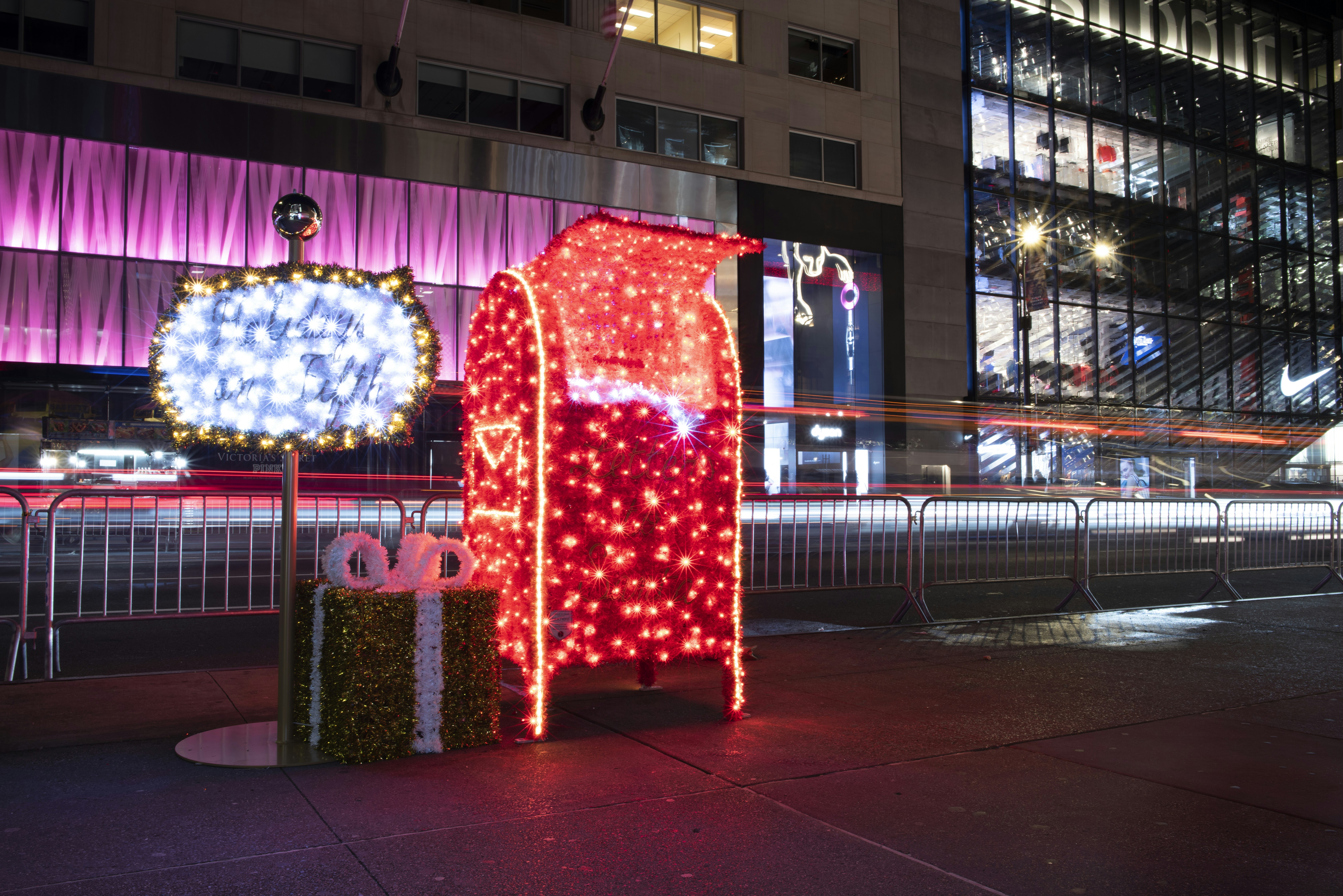 A festive mailbox adorned with colorful lights stands next to a decorated gift box, set against a backdrop of city lights and storefronts.
